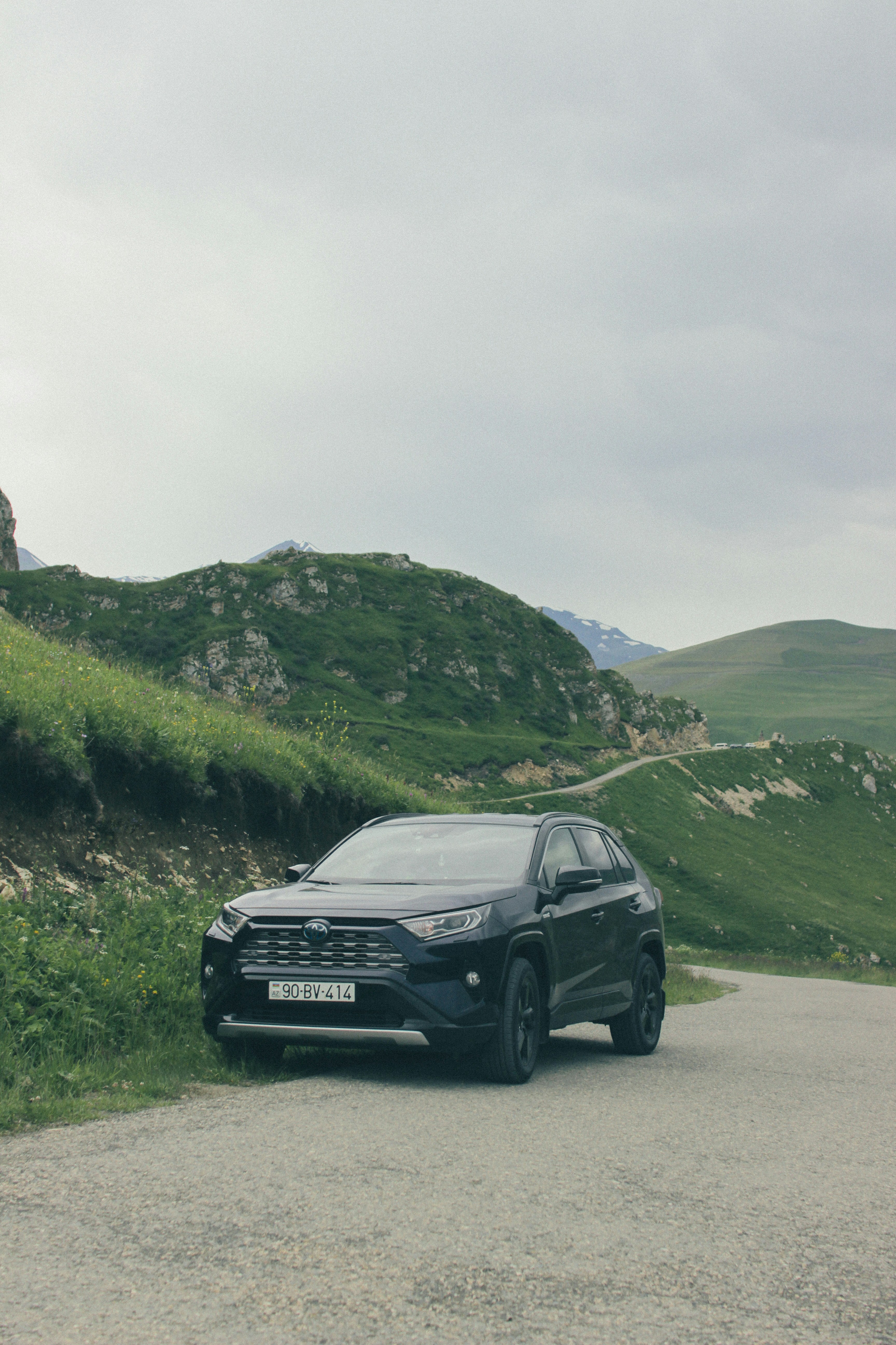 Black SUV navigating a winding road alongside lush green hills under an overcast sky.