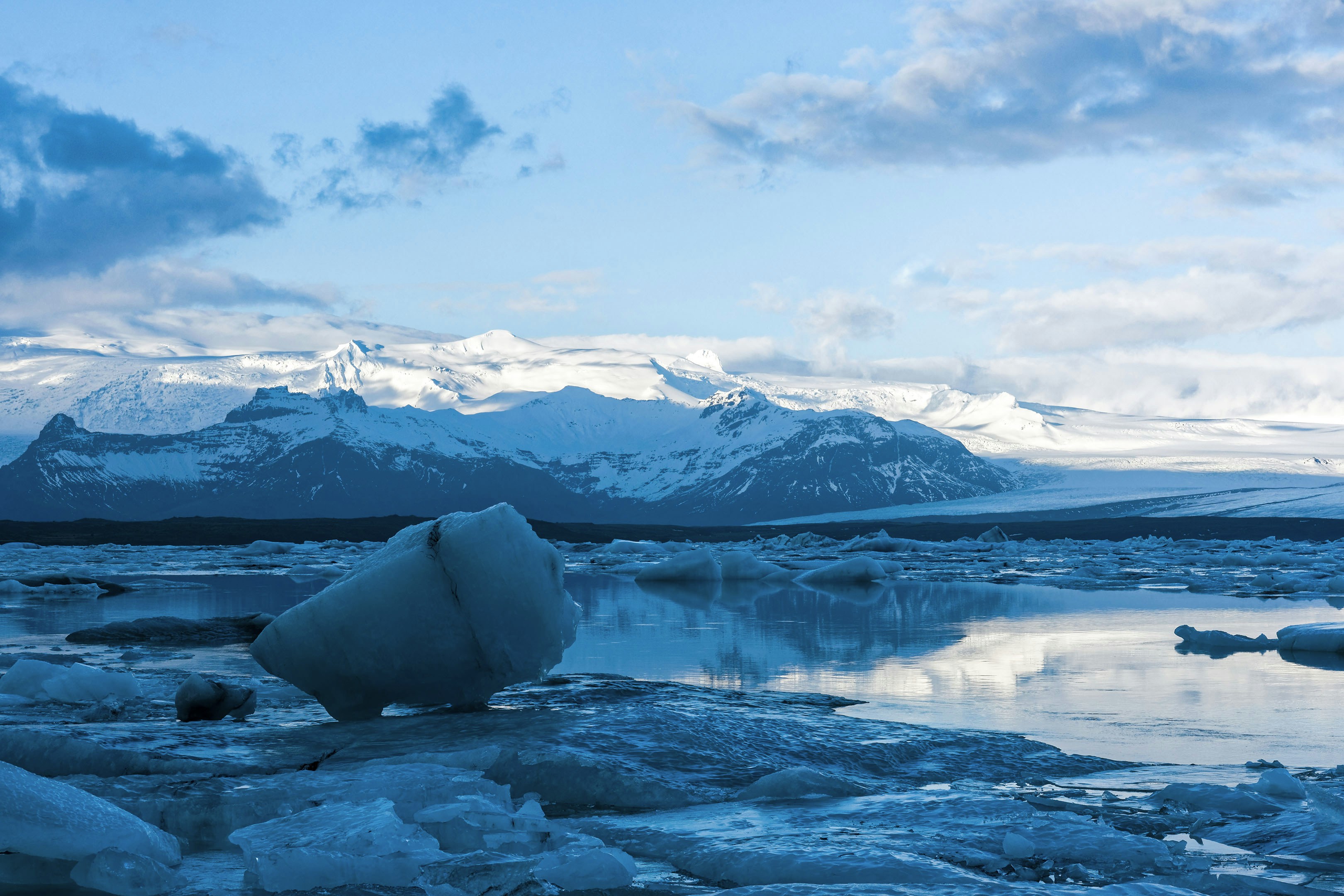 Icebergs float serenely in a blue-hued lagoon with snow-capped mountains under a partly cloudy sky.