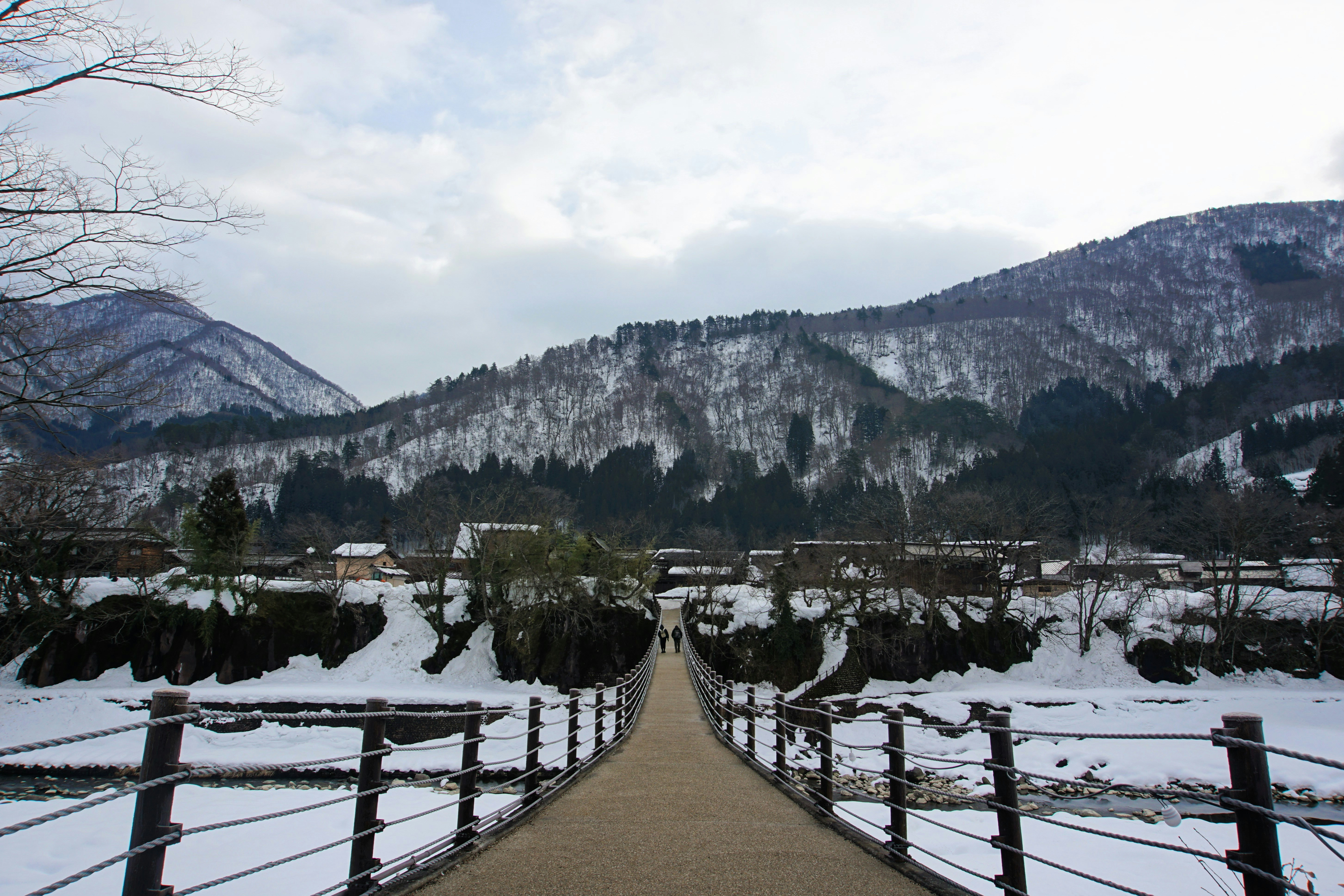 Snow-covered bridge leading to a village nestled in mountainous terrain under a cloudy sky.