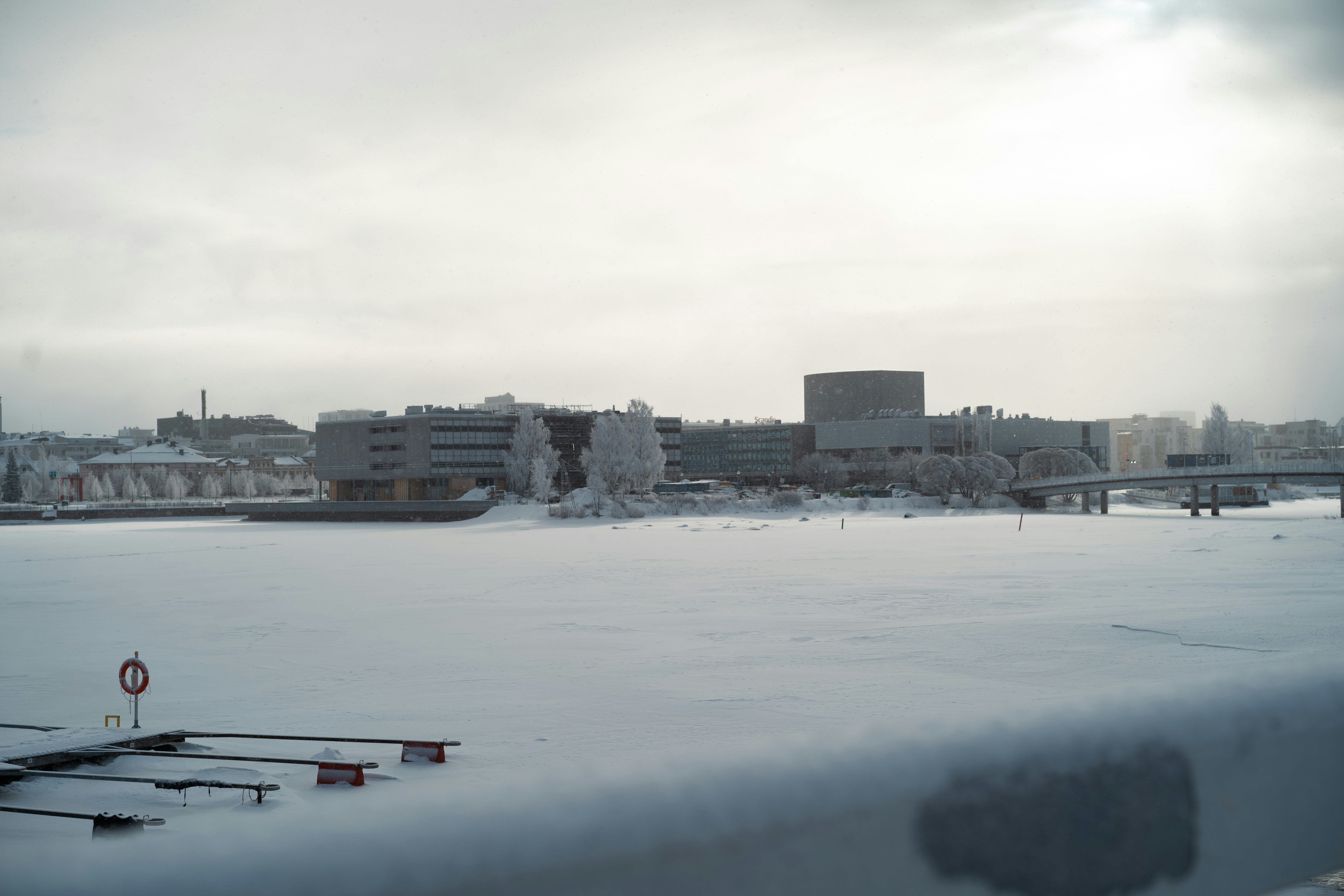 Snow-covered cityscape under a cloudy sky with a frozen river and modern buildings.