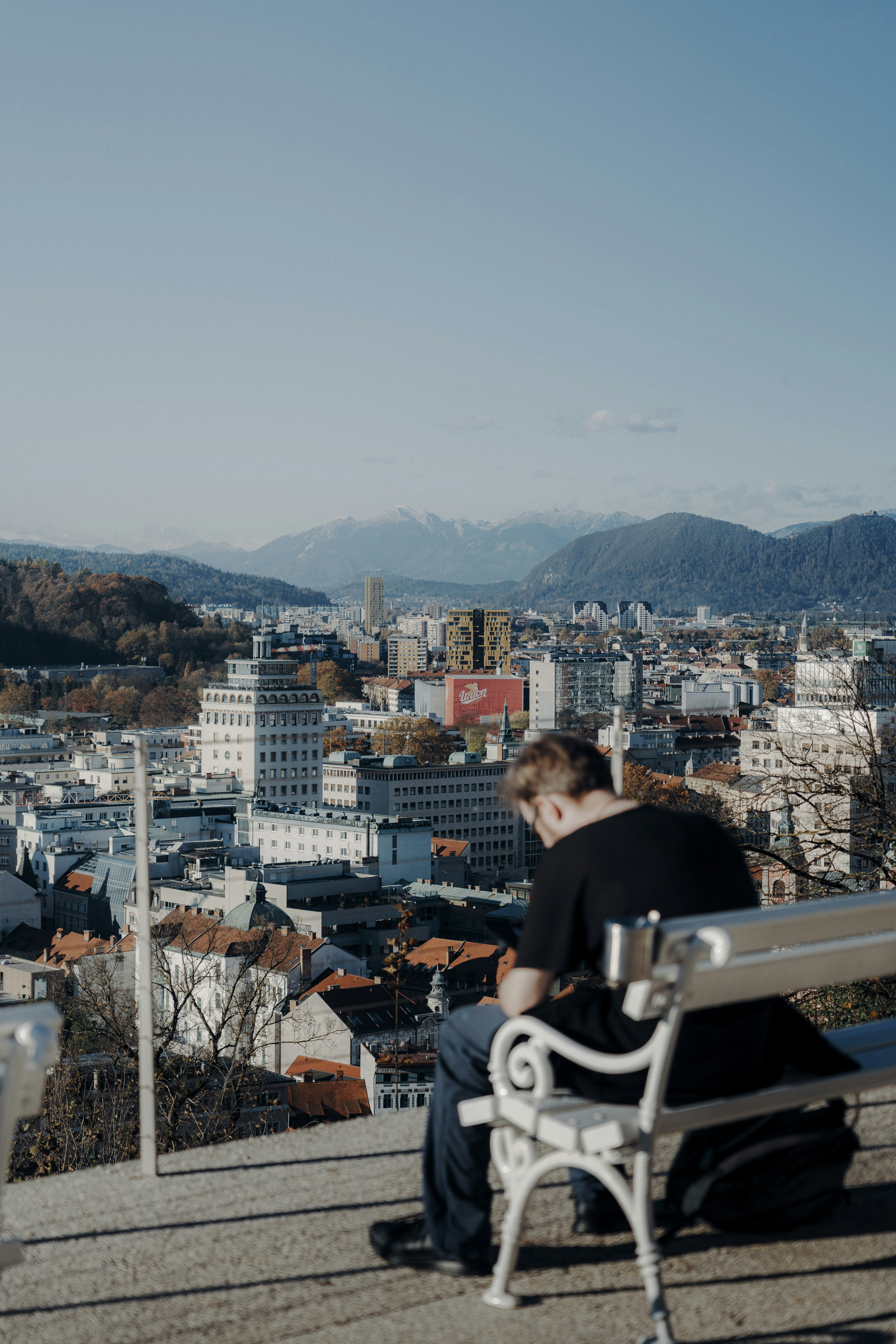 Person sitting on a bench overlooking the cityscape of Ljubljana with distant mountains under a clear blue sky.