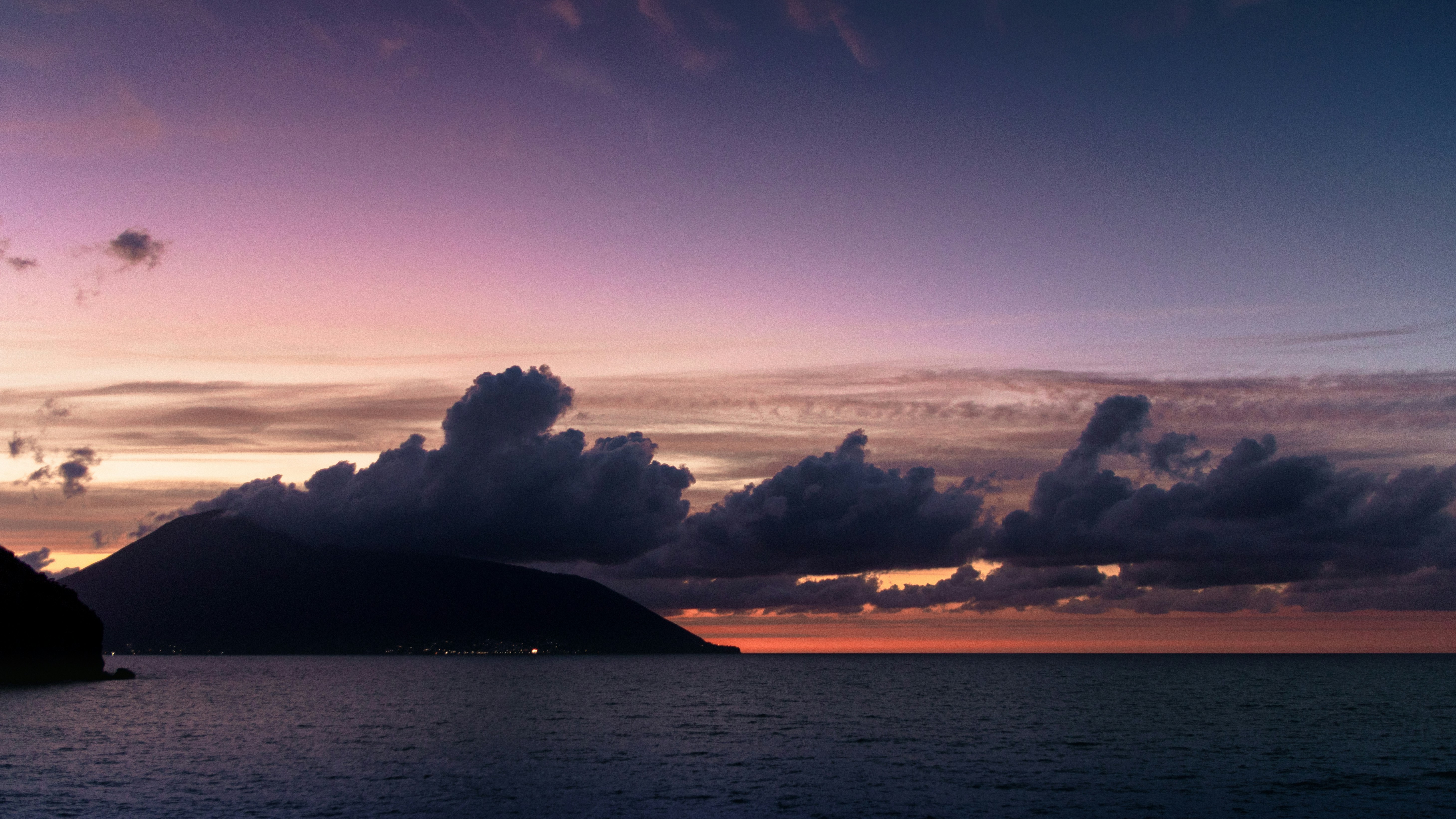 Dramatic clouds hover over a silhouetted island at sunset, casting vibrant hues across the sky.