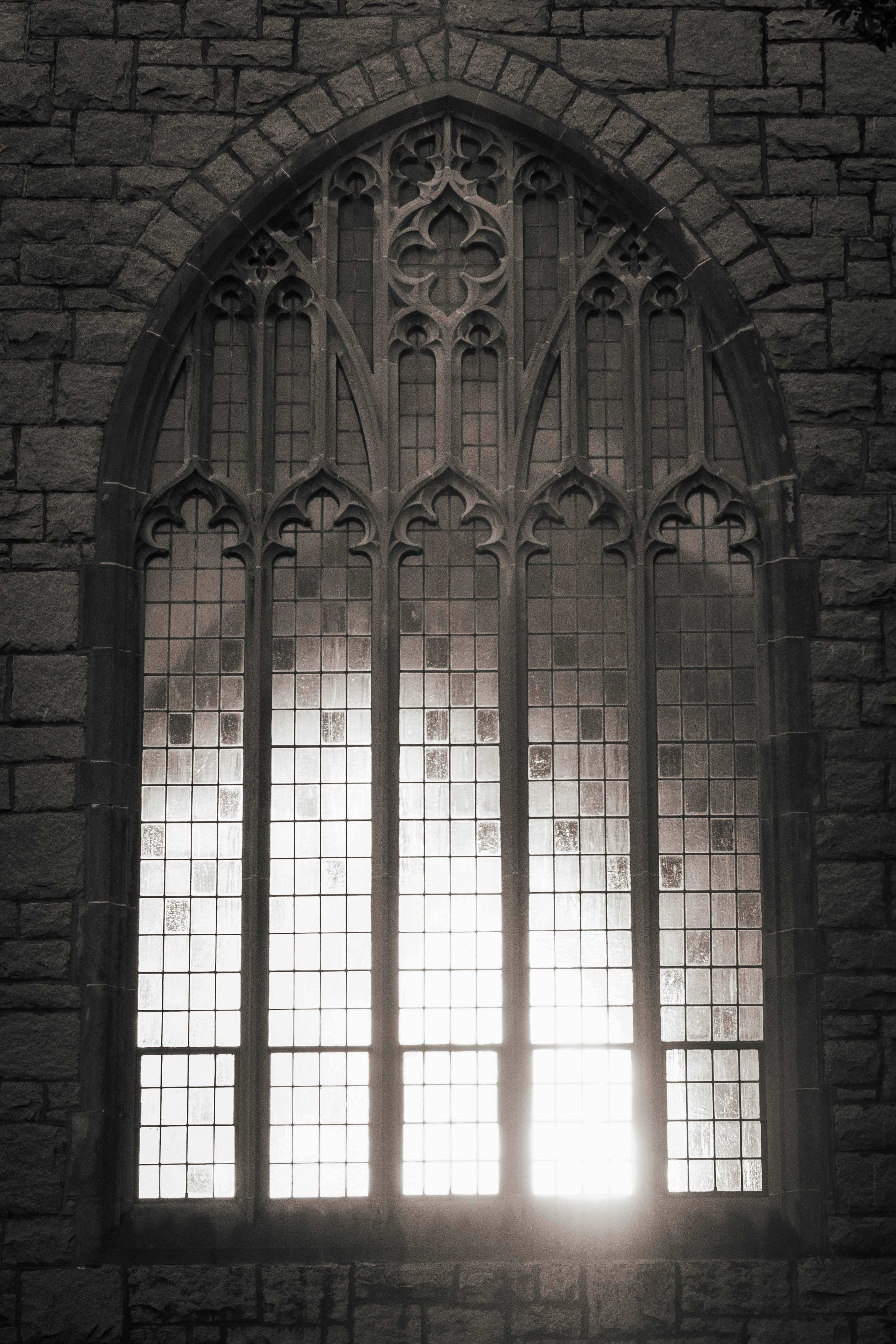 Intricate gothic window framed by textured stone wall, illuminated by soft light from outside.