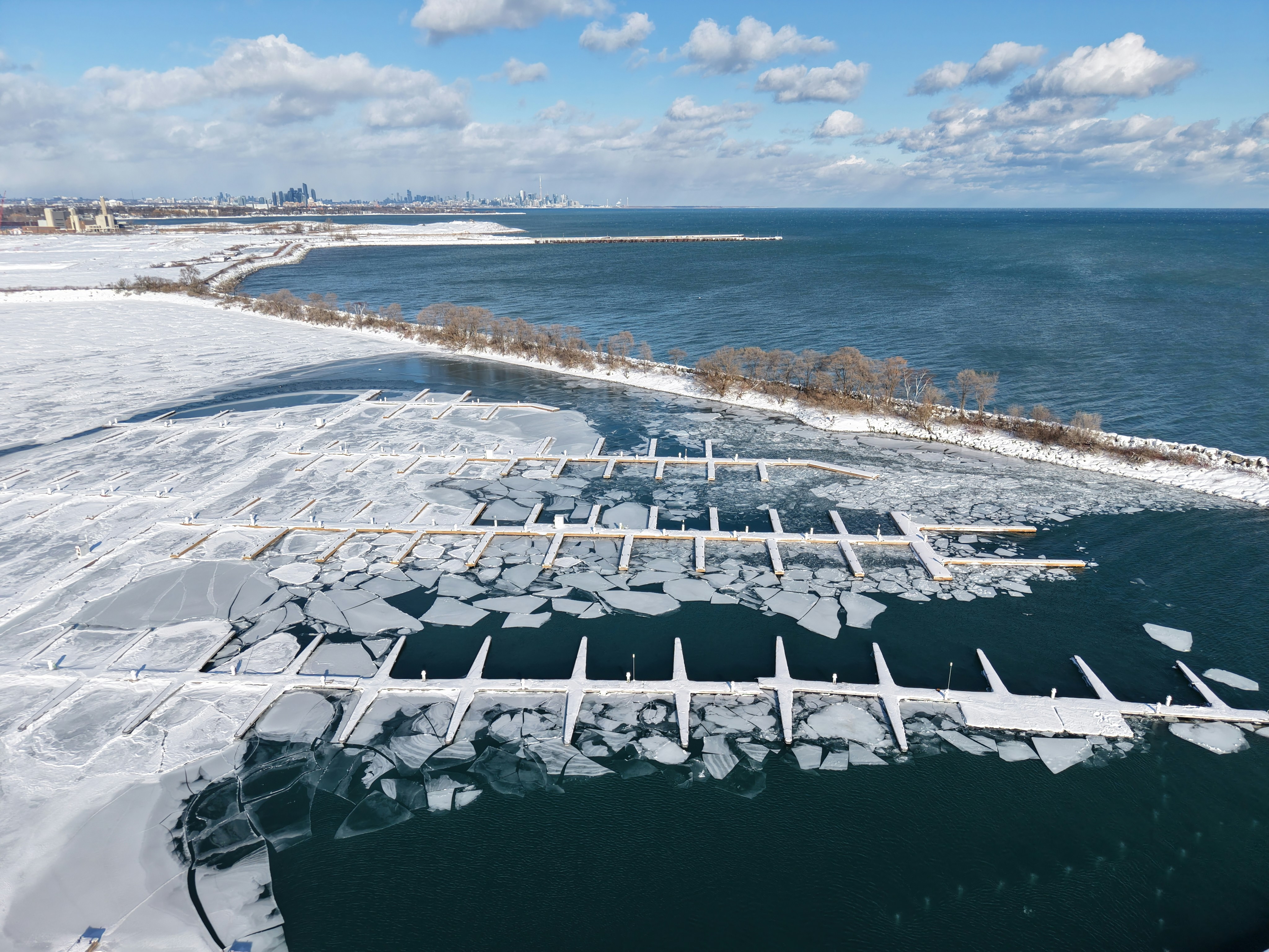 Aerial view of a marina partially frozen over, with ice formations contrasting against the deep blue water and distant city skyline. 