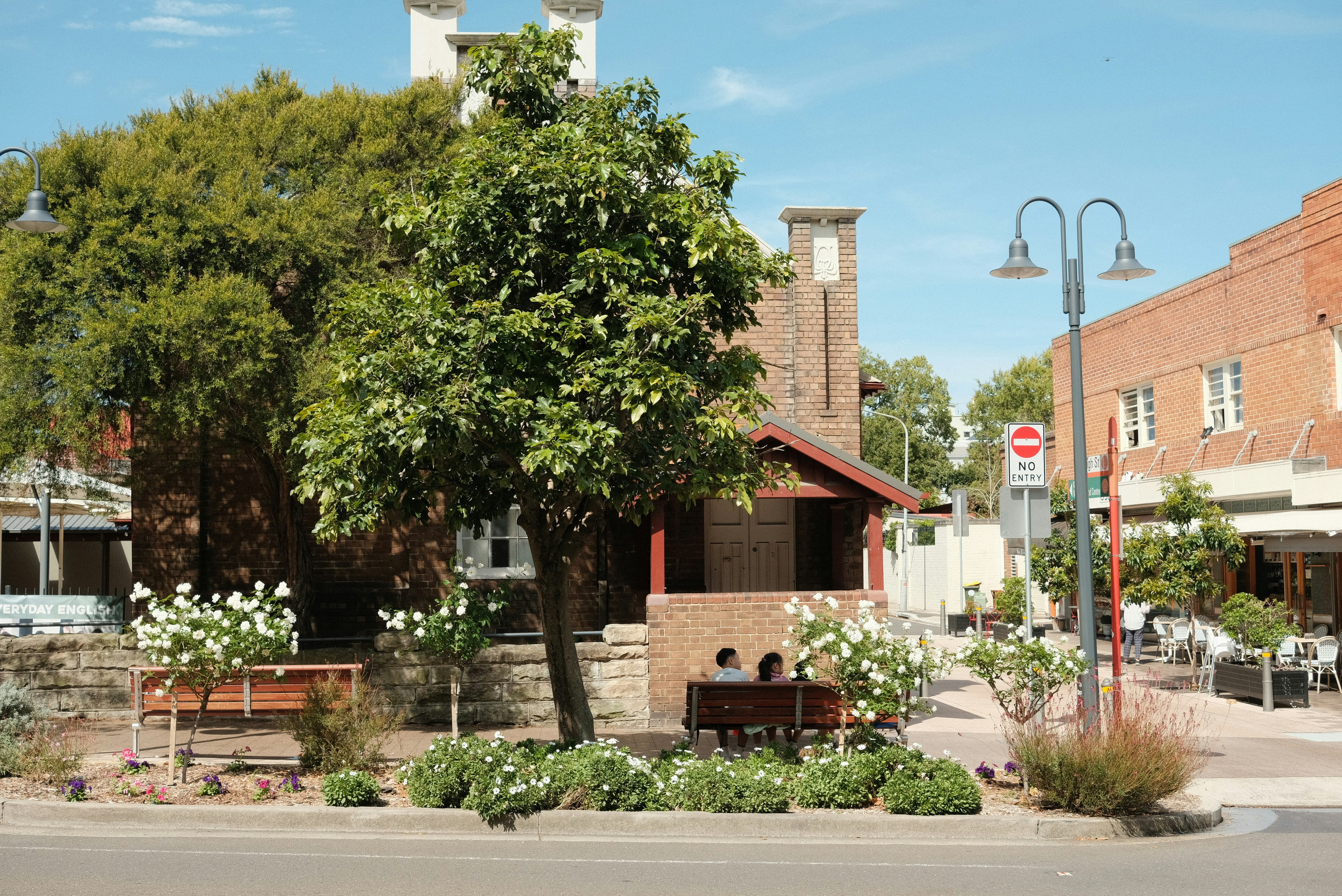 A tree-shaded bench near a small brick building and clock tower, surrounded by white flowers and urban architecture.
