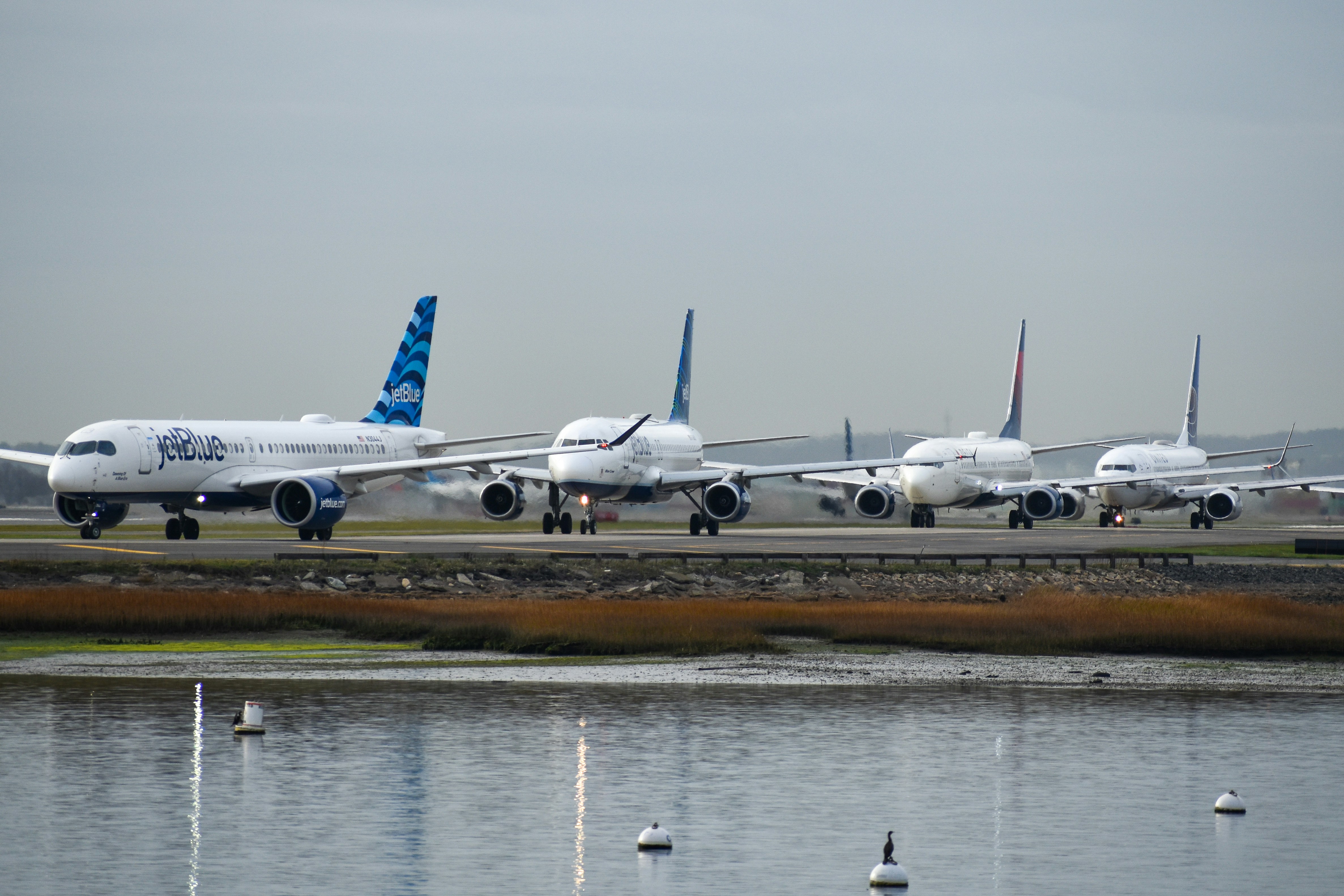 Airplanes queued on a runway near a reflective water surface, ready for takeoff.