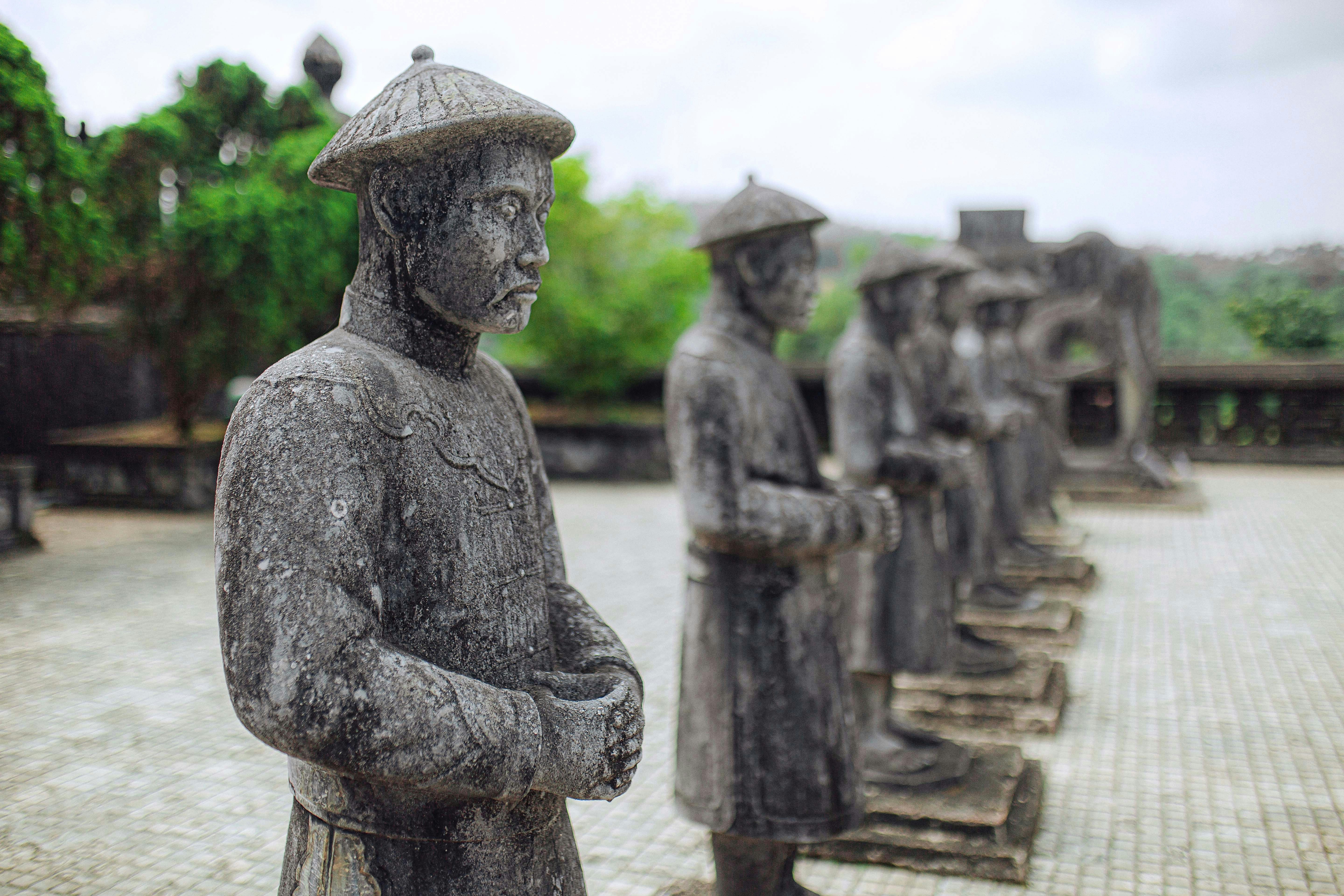Stone statues of soldiers in a line, set against lush greenery and a cloudy sky.