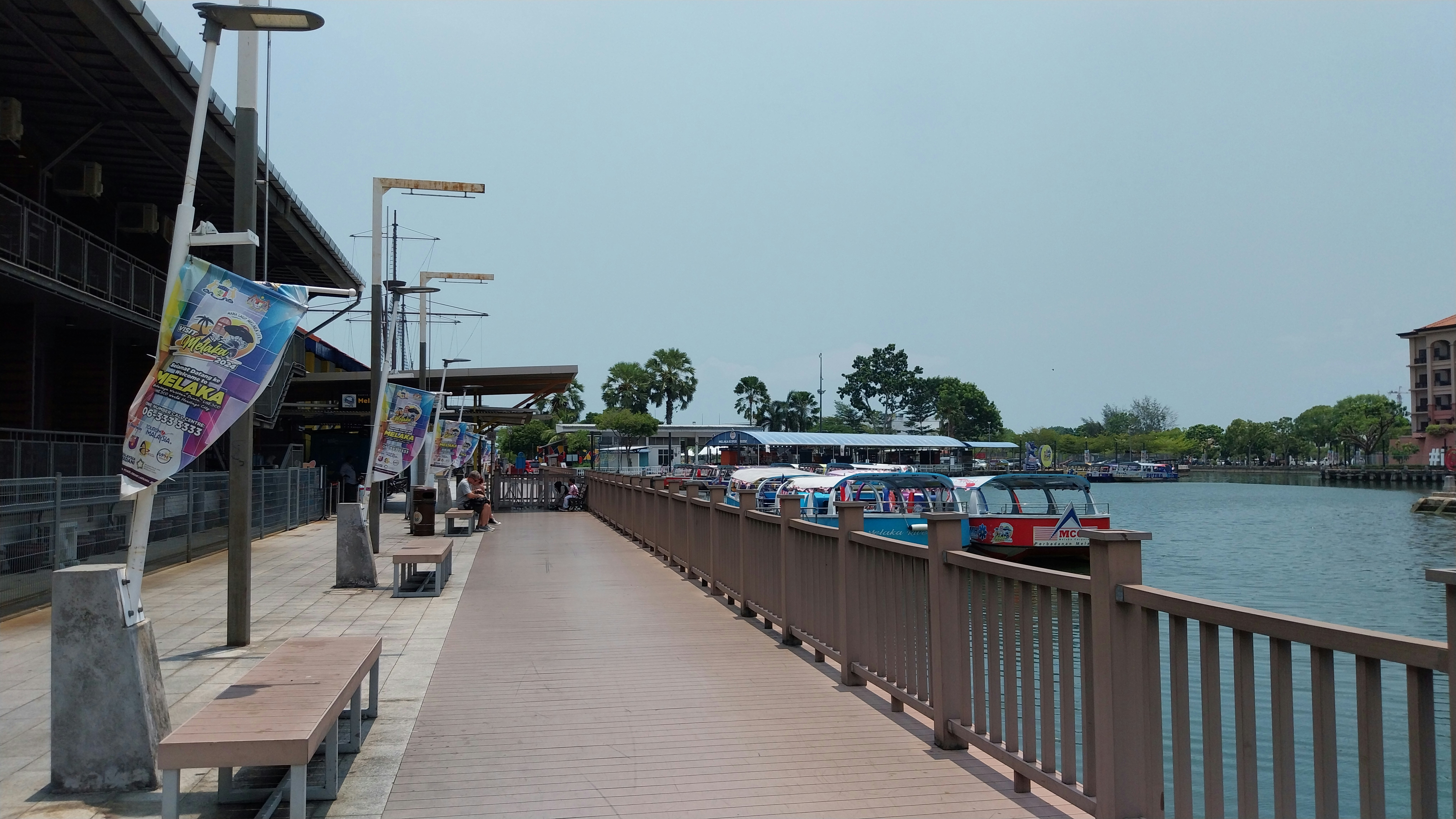 Pedestrian walkway alongside a river with moored boats and distant trees on a clear day.