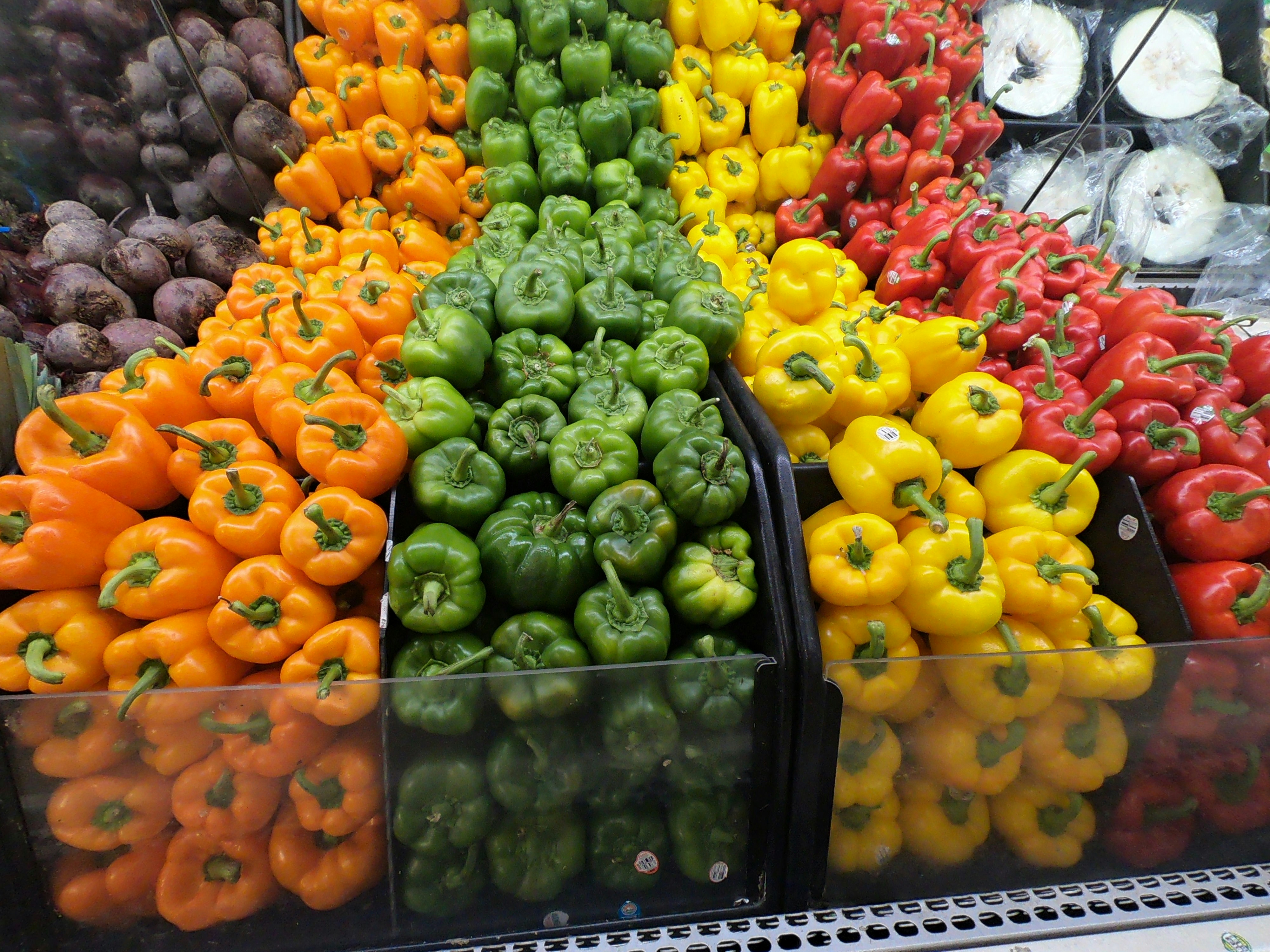 Colorful bell peppers are neatly stacked in crates by hue at a market, forming rainbow rows. The arrangement highlights oranges, greens, yellows, and reds with onions visible in the background.