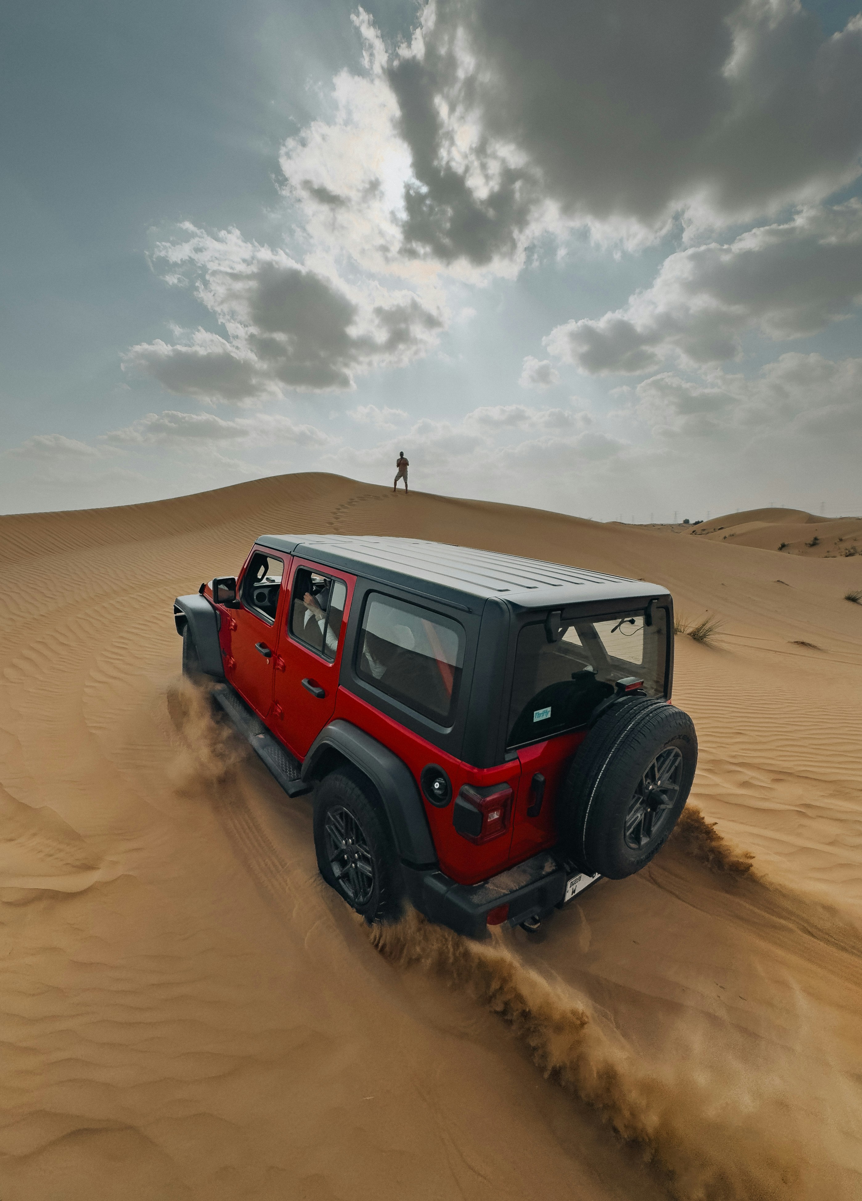 Red off-road vehicle navigating sandy dunes under a dramatic sky, with a lone figure in the distance. Dust trails swirl around the tires.