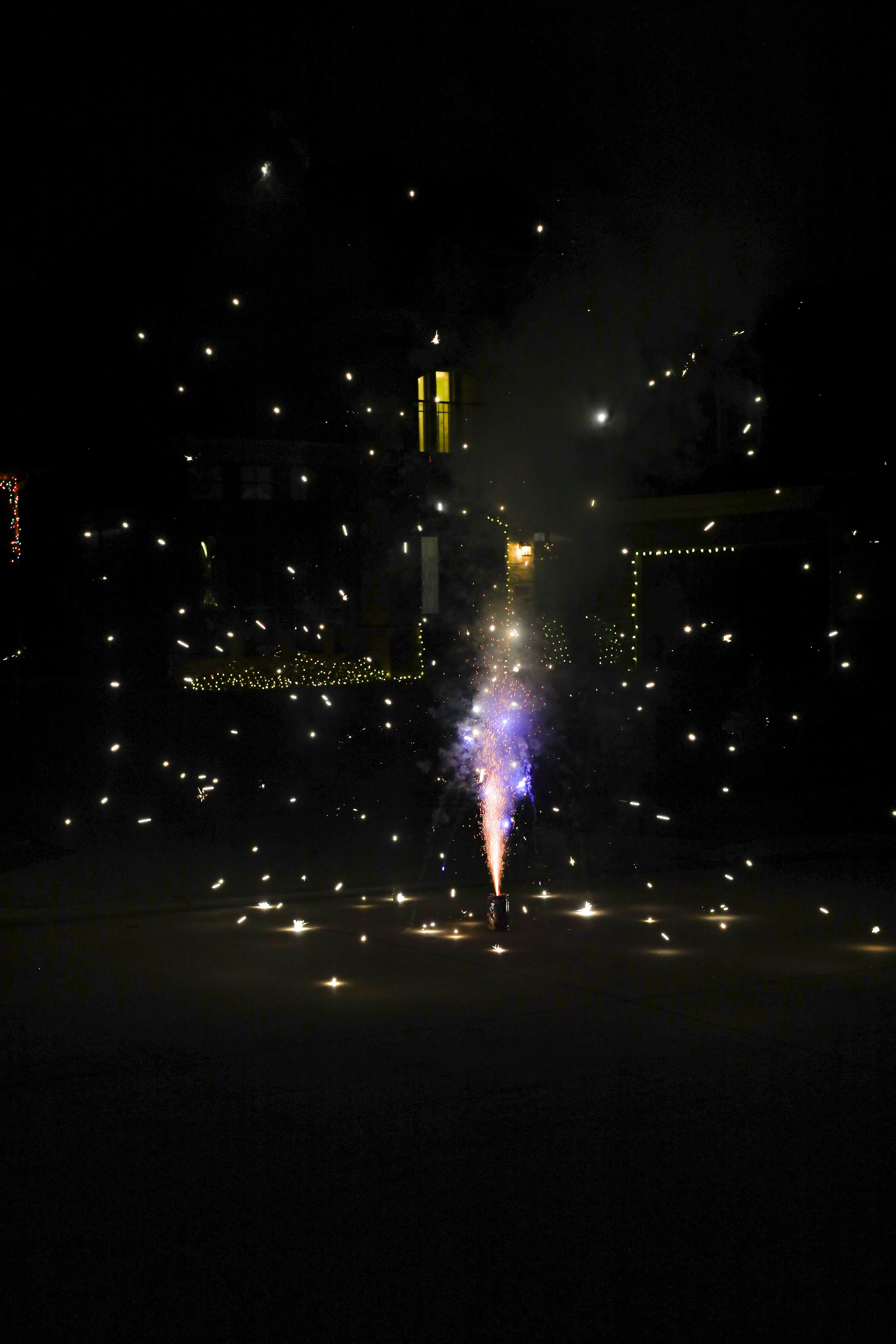 Colorful fountain firework erupting in a darkened street, surrounded by scattered sparks and illuminated holiday lights in the background.