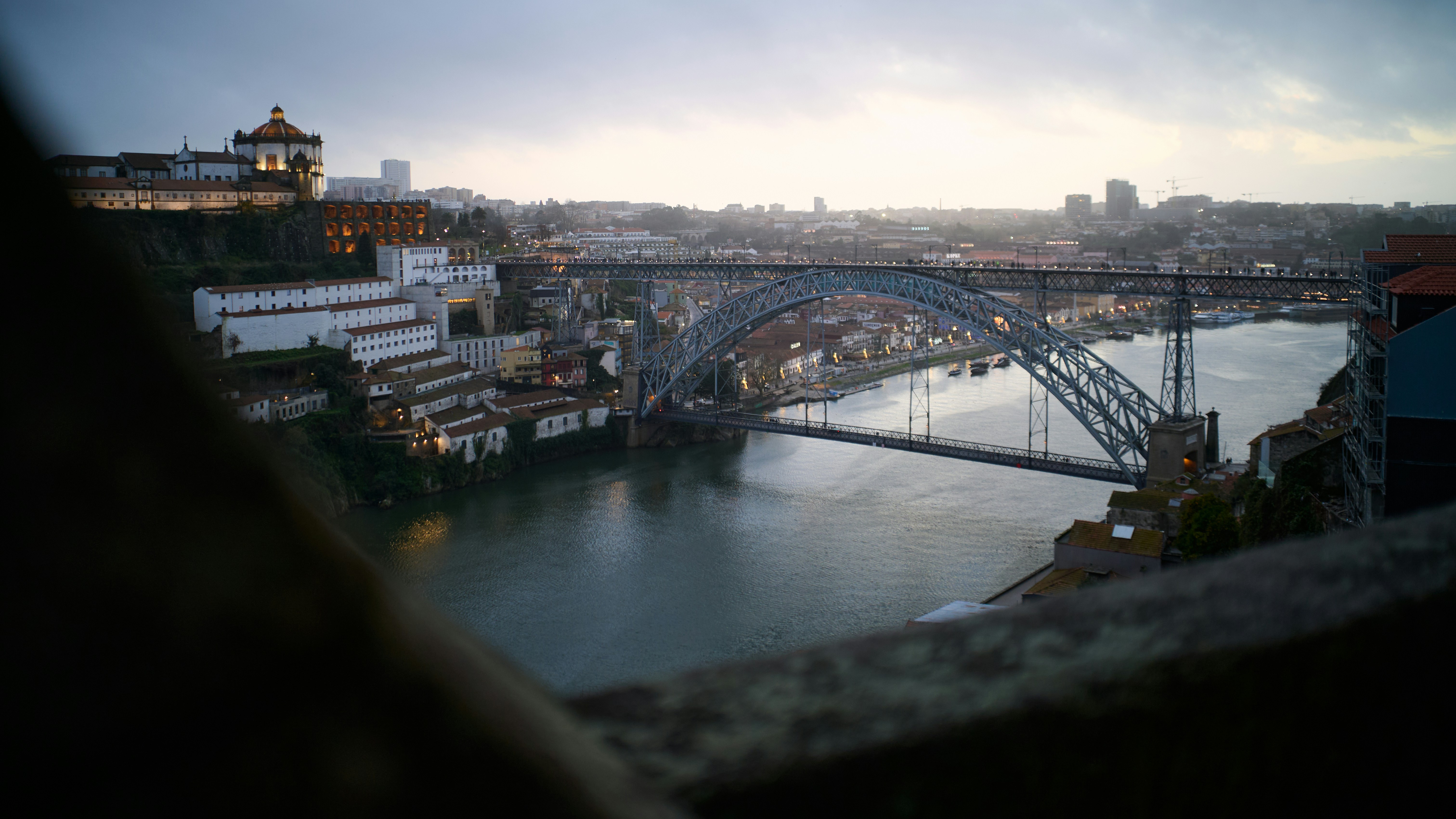 Steel arch bridge spans a serene river, with city lights glowing under a cloudy sky.