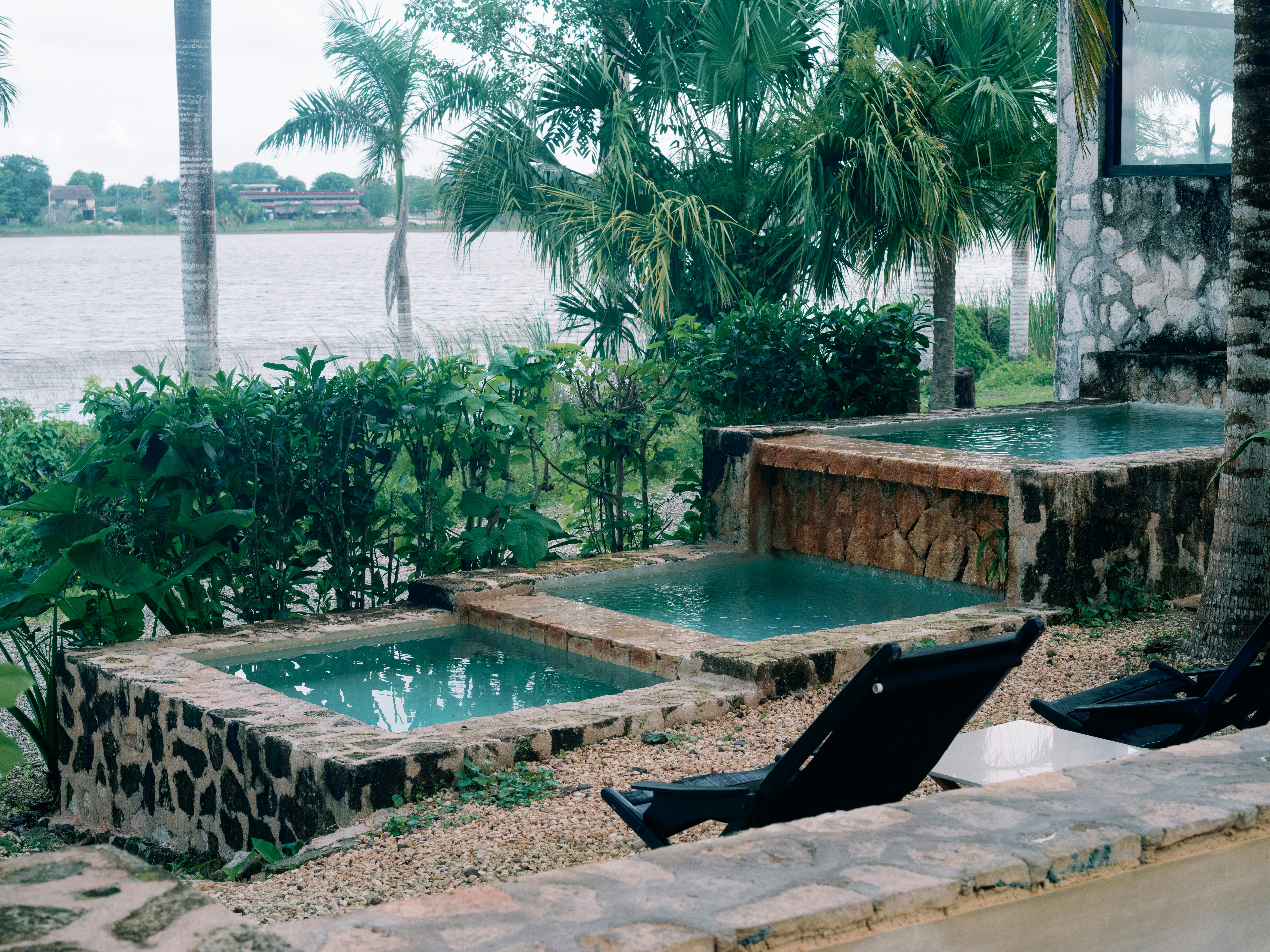 Terraced stone pools surrounded by lush greenery overlooking a serene river under a cloudy sky.