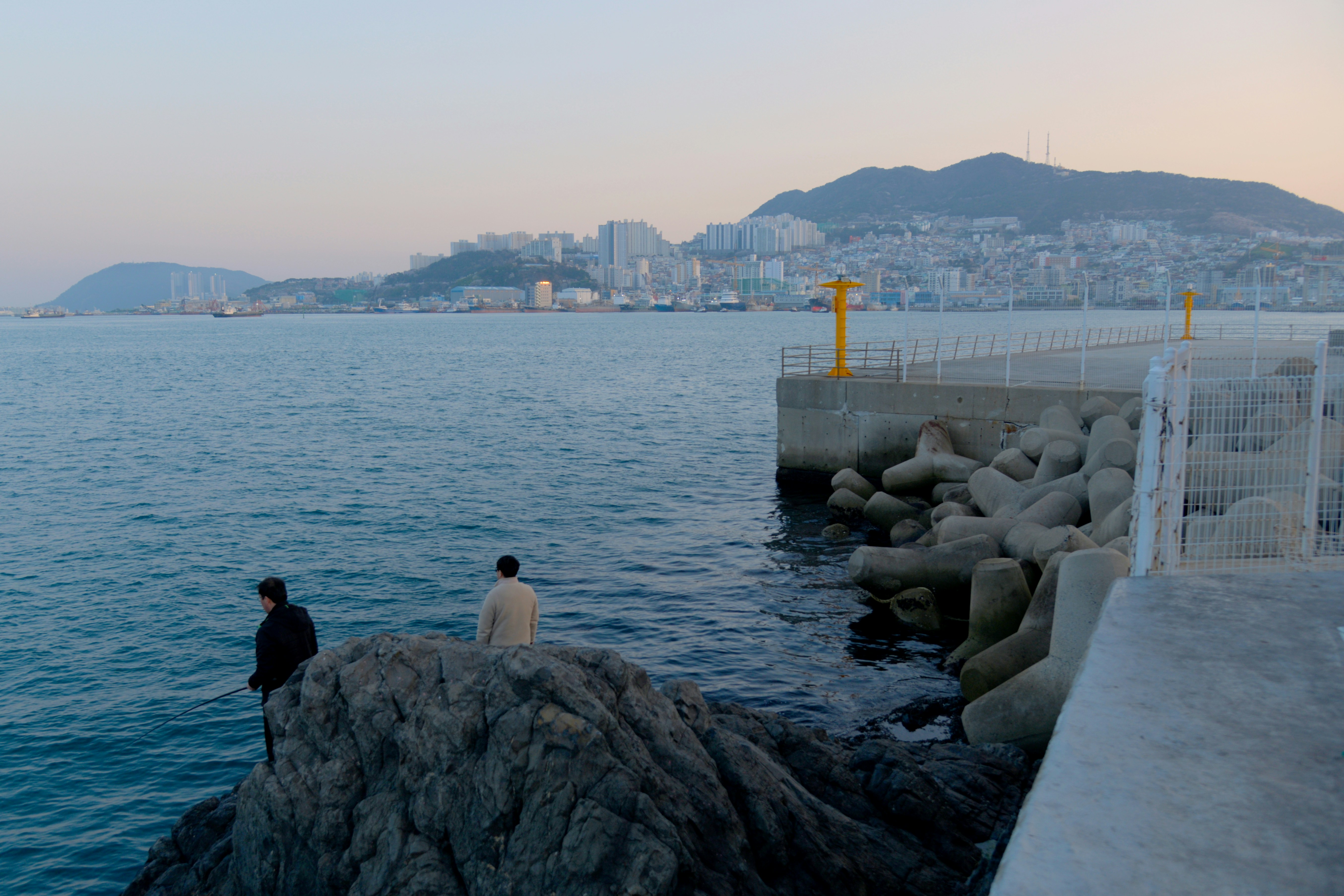Blue Hour at the Breakwater: Two Figures on Worn Rocks with a Distant City