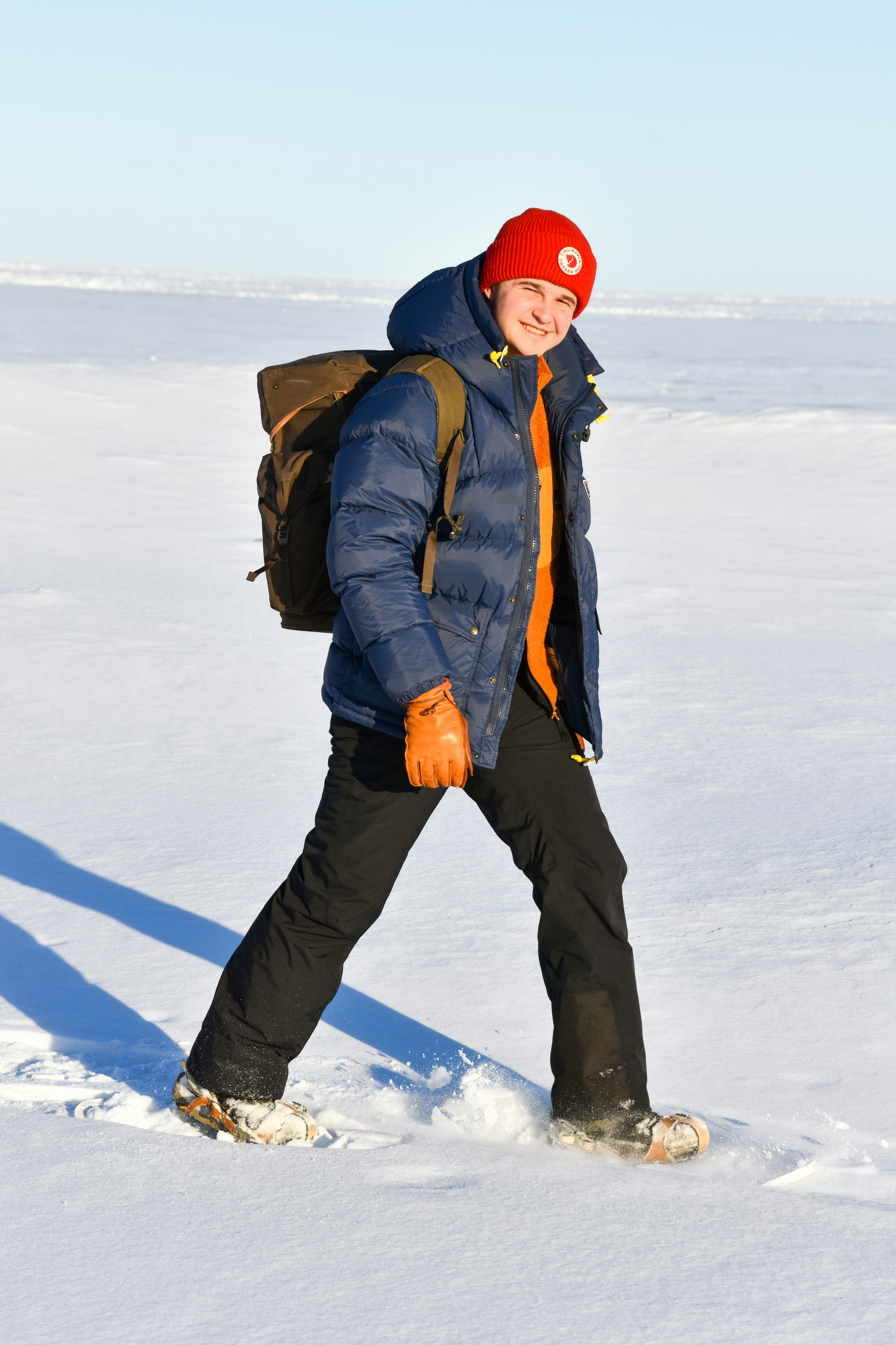 Person in winter gear walking through a sunlit snowfield with a backpack and red hat.