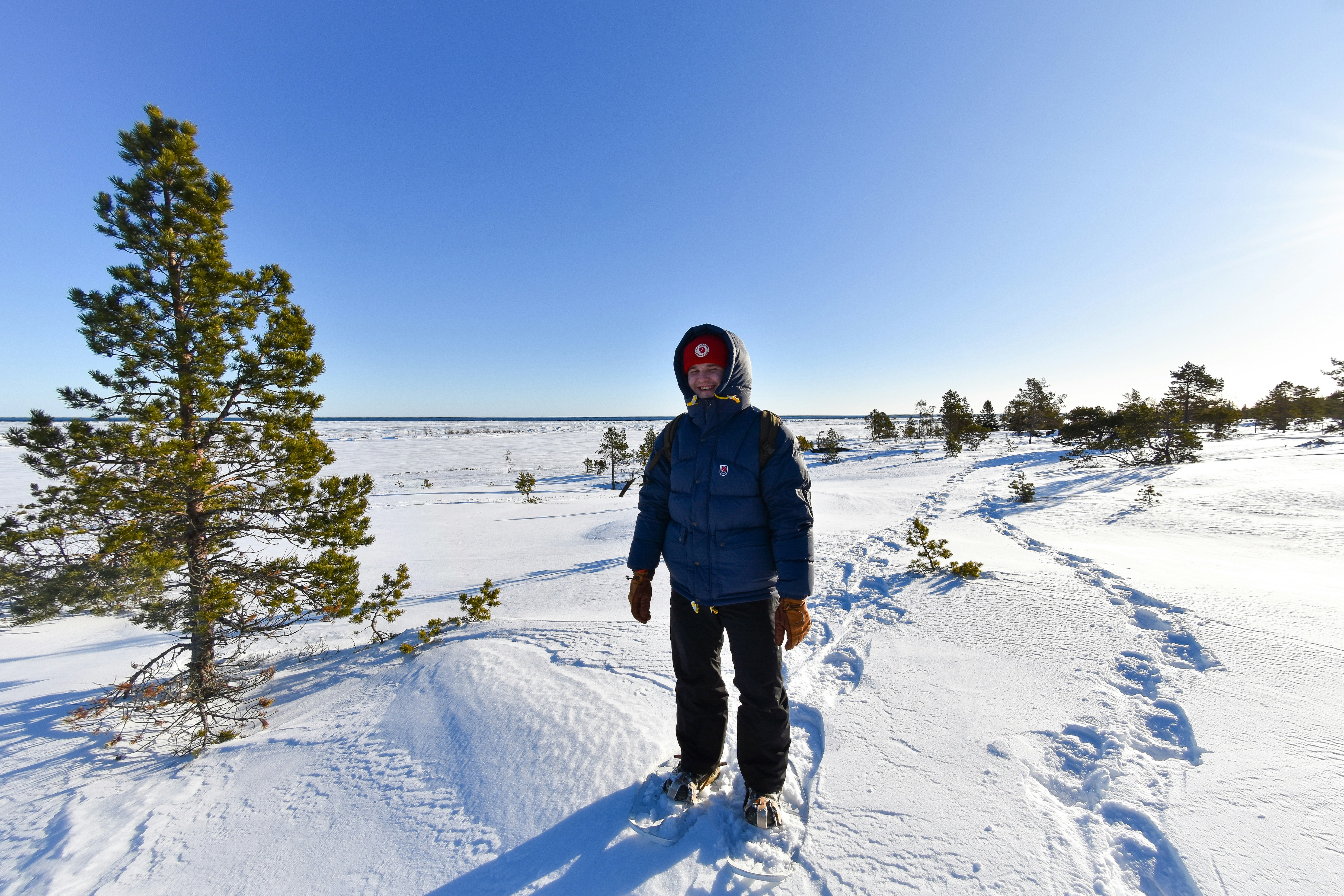 Person in snowshoes stands on a snowy landscape with scattered trees and clear blue sky.