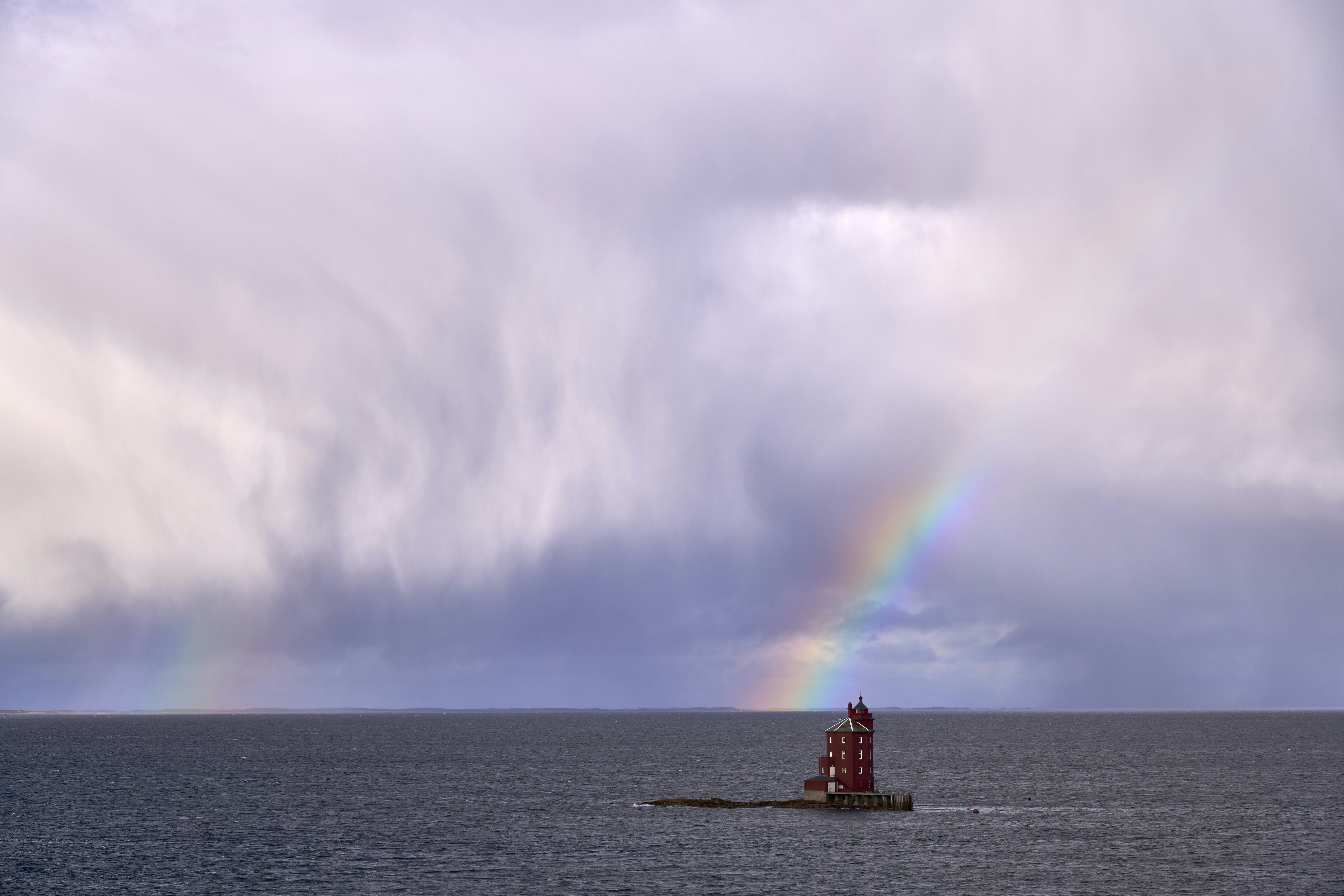 Lonely lighthouse stands against a vast ocean backdrop with a vibrant rainbow arching through stormy skies.