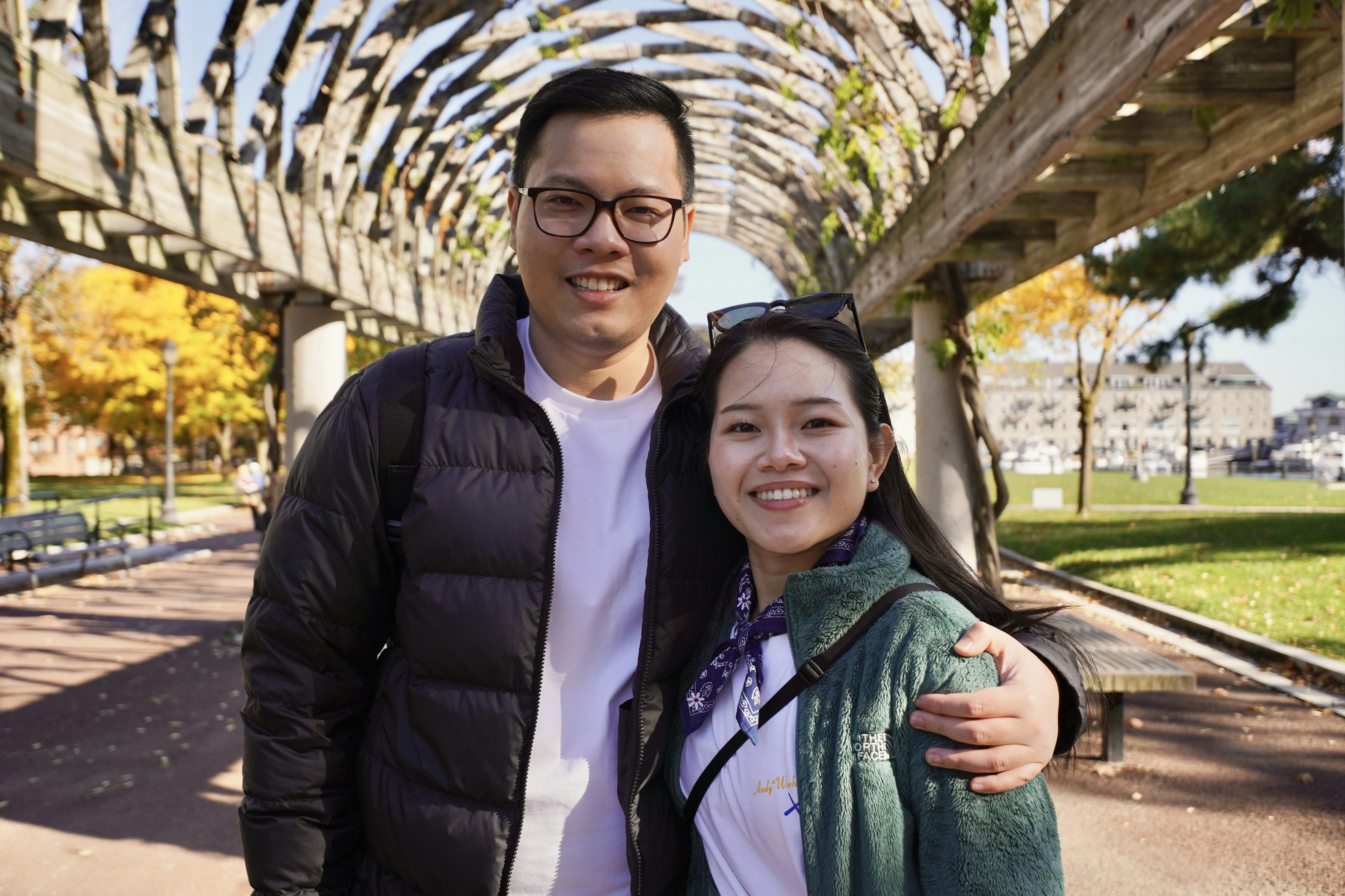 Smiling couple under an arched pergola in a sunlit park with golden autumn leaves.