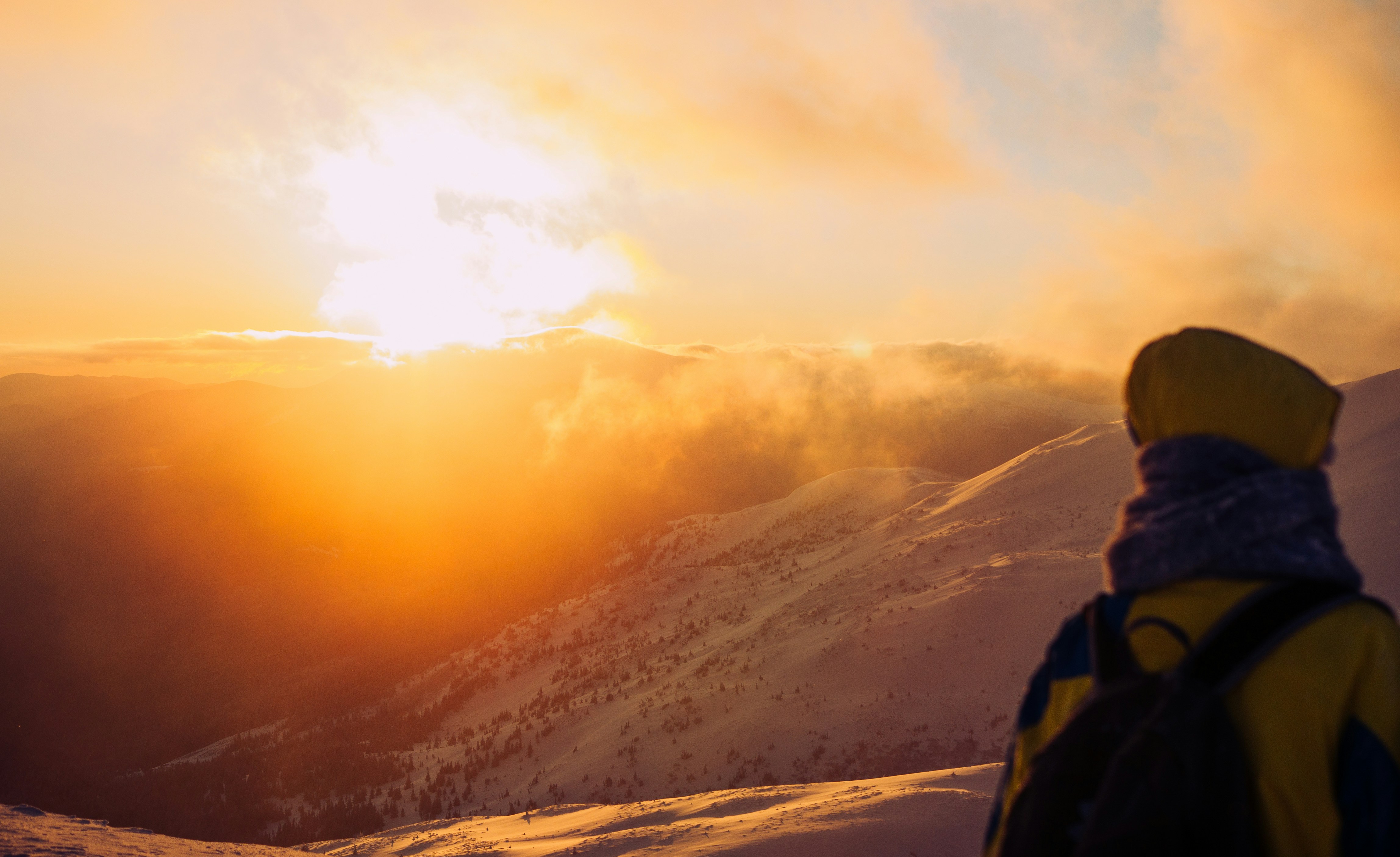Person in winter attire gazing at a vibrant sunrise over snow-covered mountains.