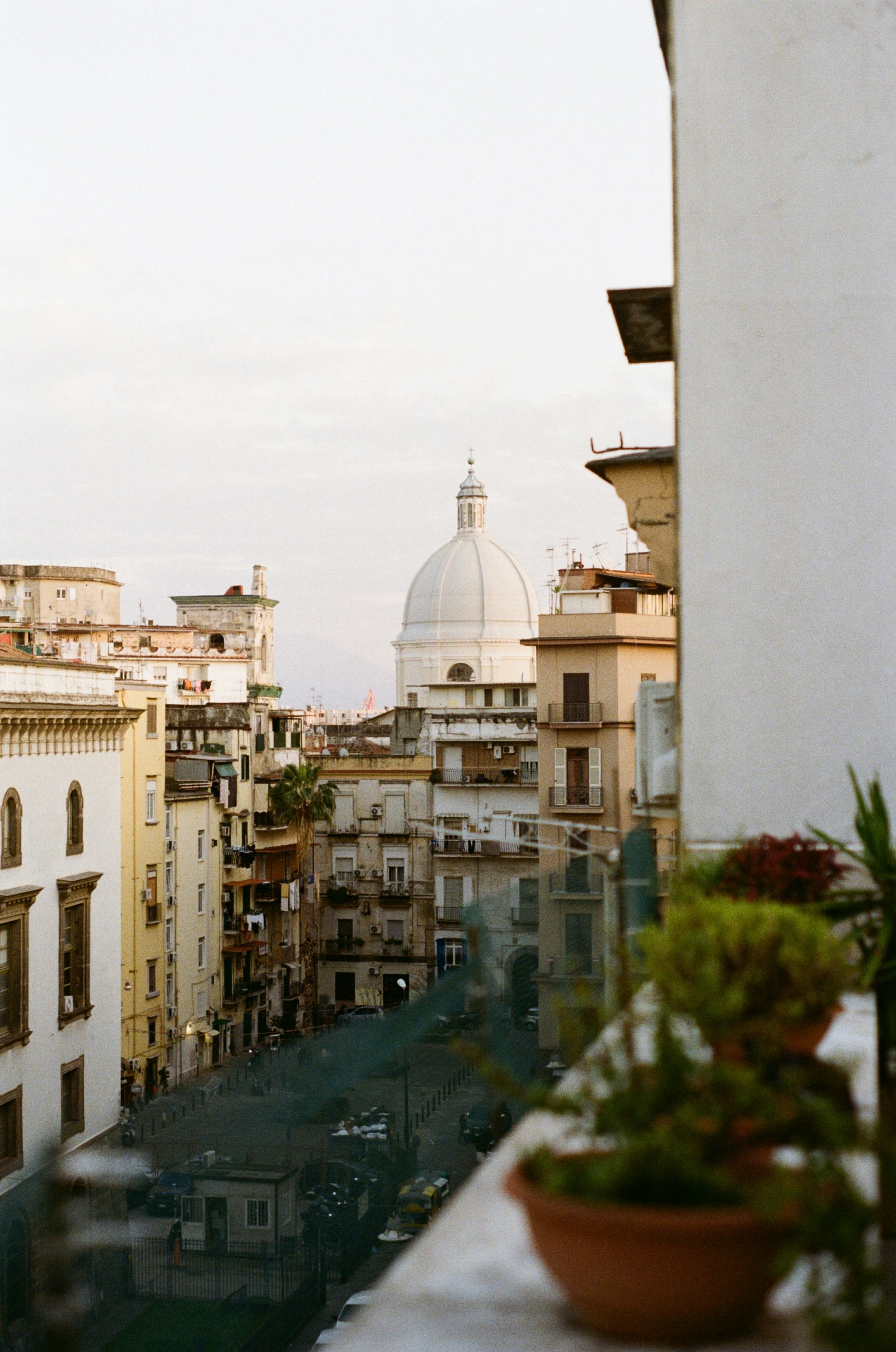 City street scene framed by a balcony railing, with a domed building rising above the rooftops.