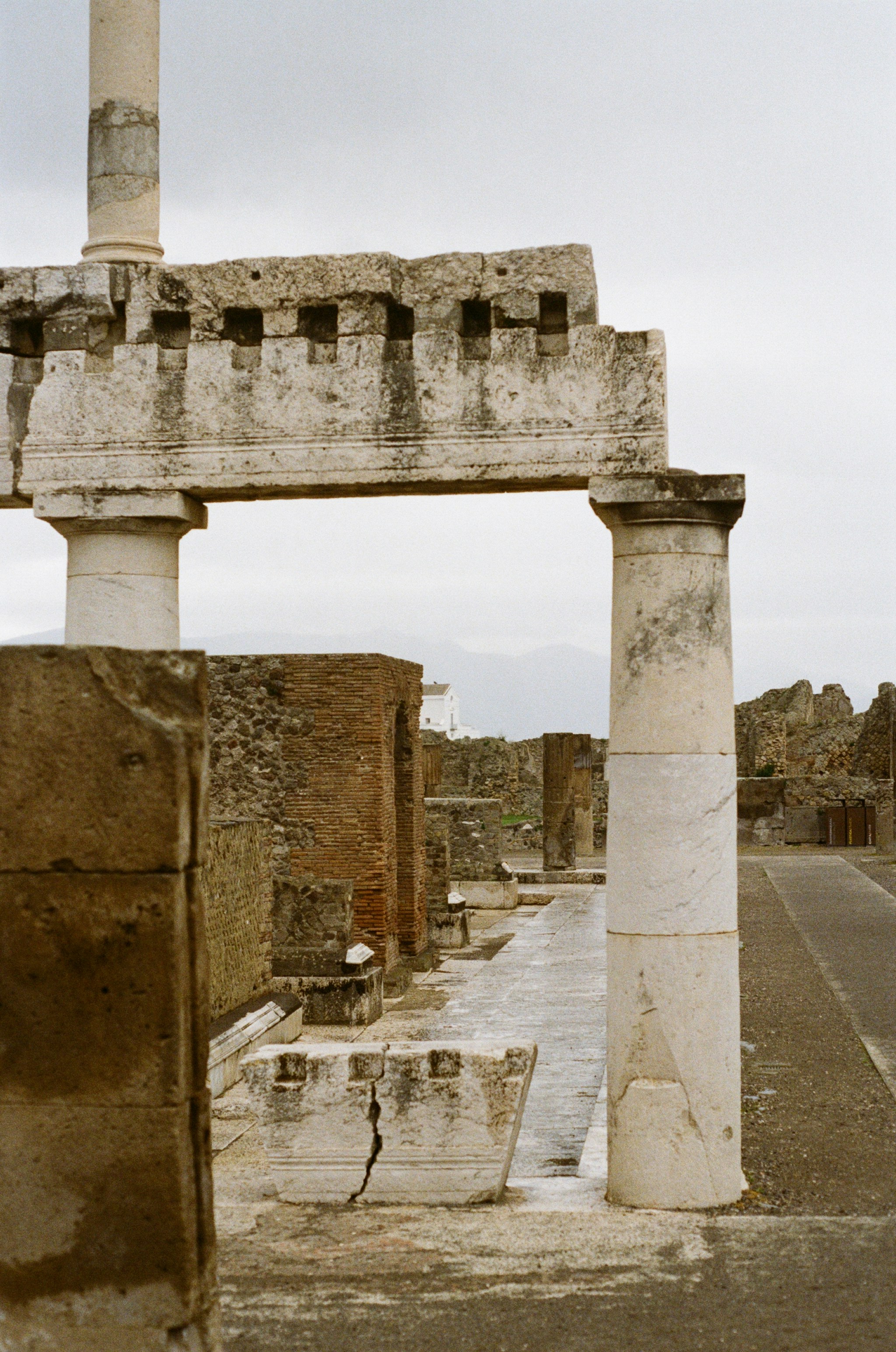 Ancient ruin featuring weathered columns and a stone arch that frames a narrow corridor of brick walls, leading toward hazy mountains under a gray sky.