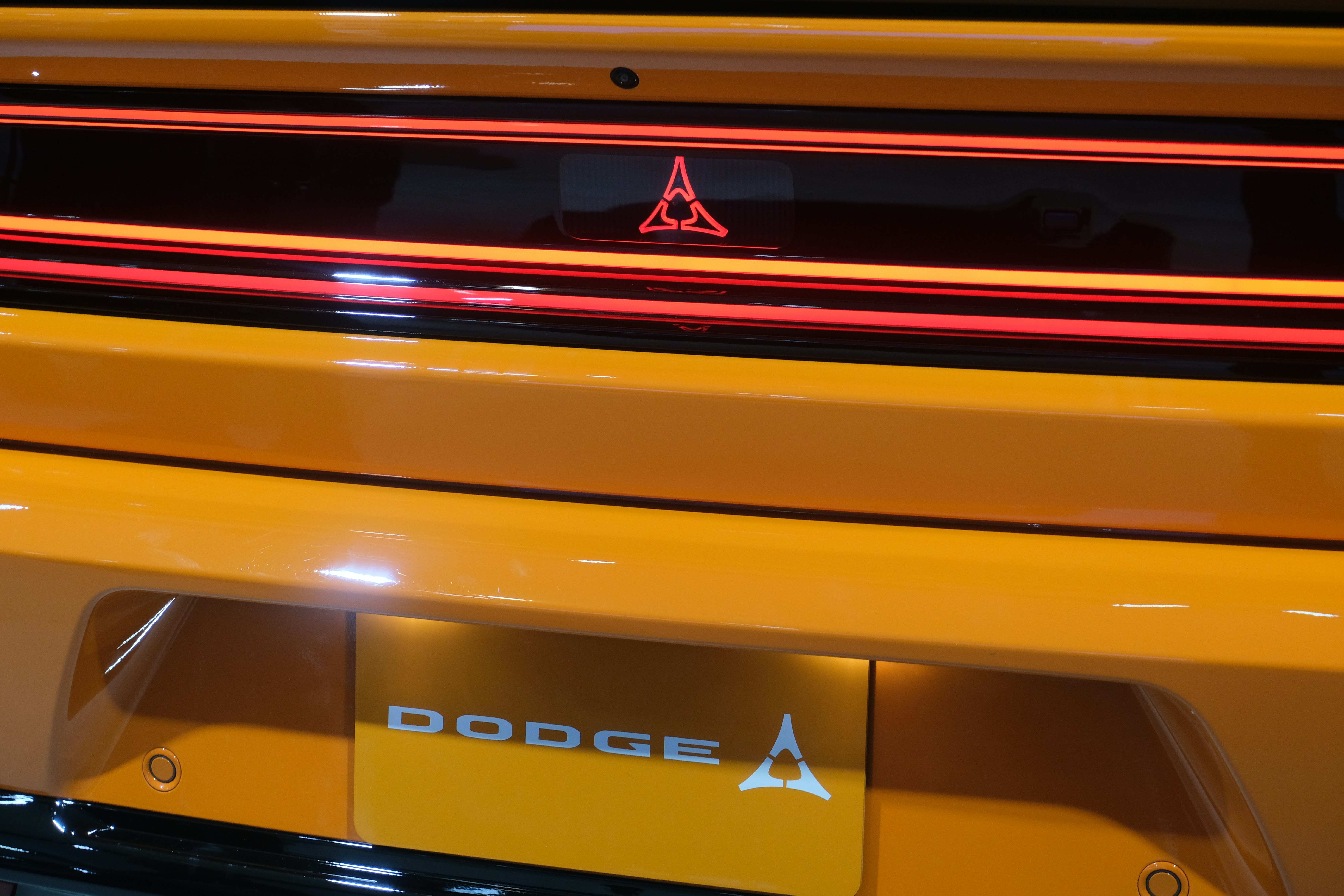 Close-up of a Dodge vehicle's rear, showcasing the illuminated logo and sleek design elements against a bold orange background.