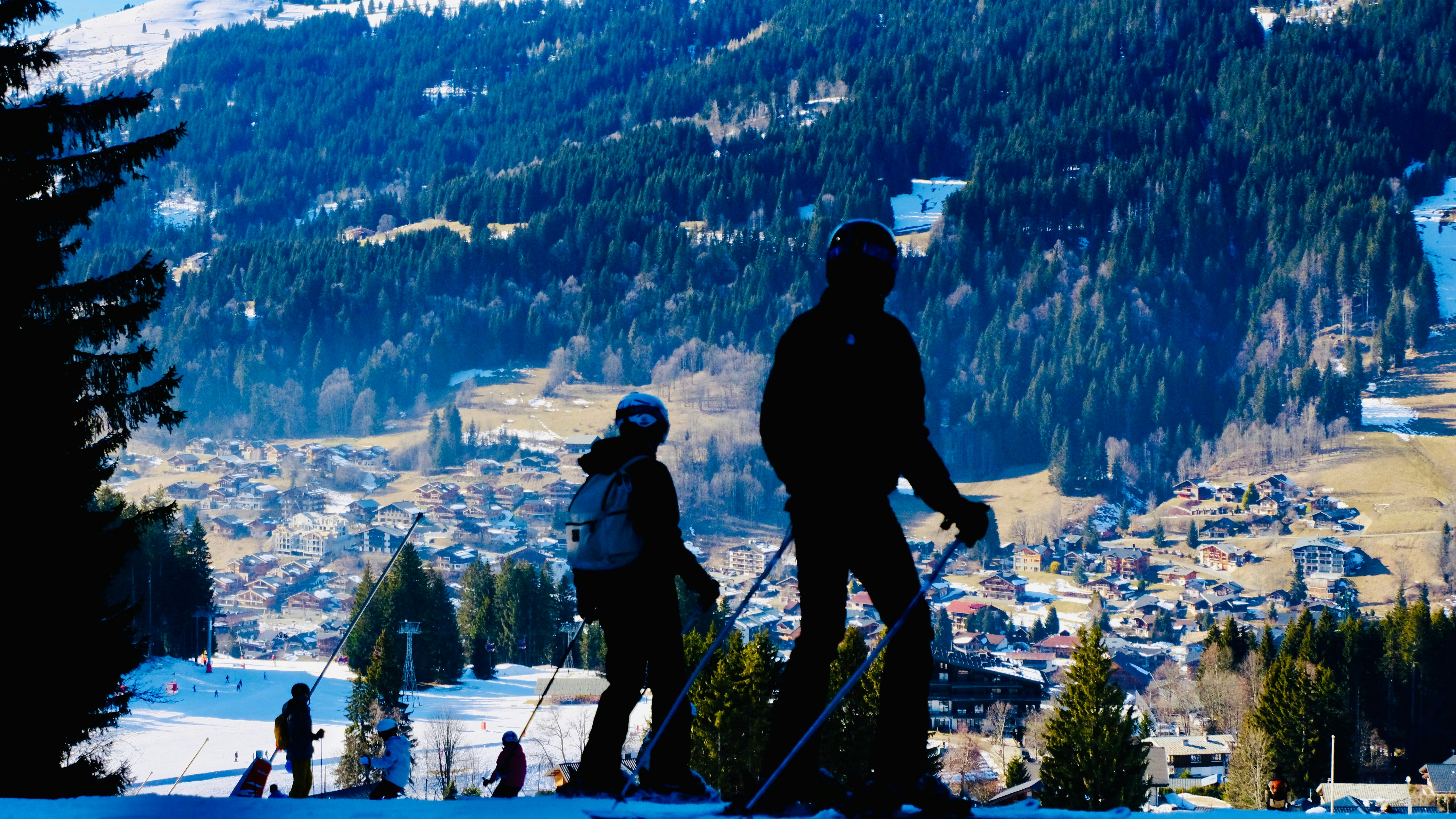 Skiers navigating a snowy slope against a backdrop of a picturesque alpine village nestled in the valley. The silhouettes create a striking contrast with the vibrant landscape.