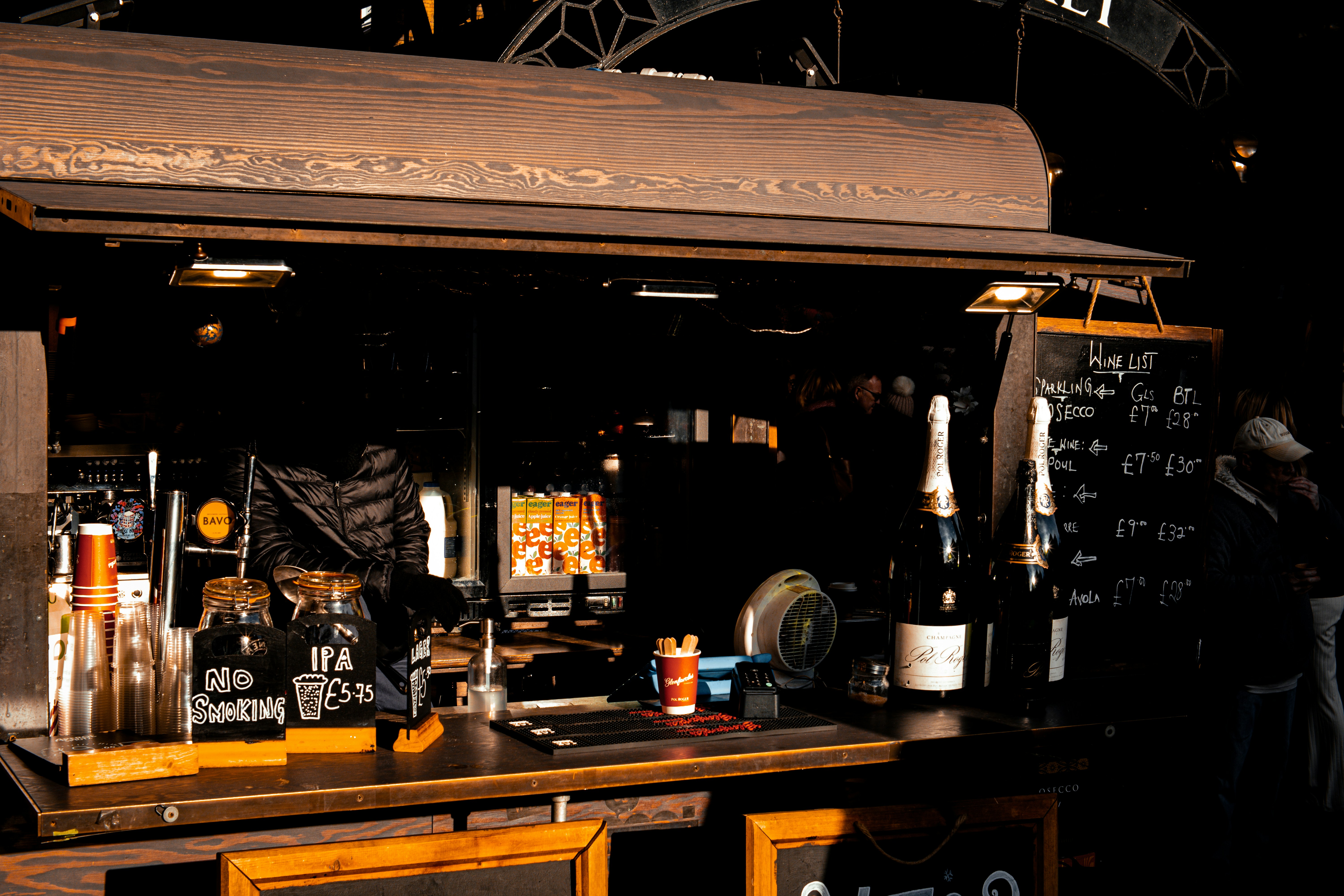 Market stall with beverages and pastries under warm lighting, featuring a chalkboard menu and large champagne bottles.
