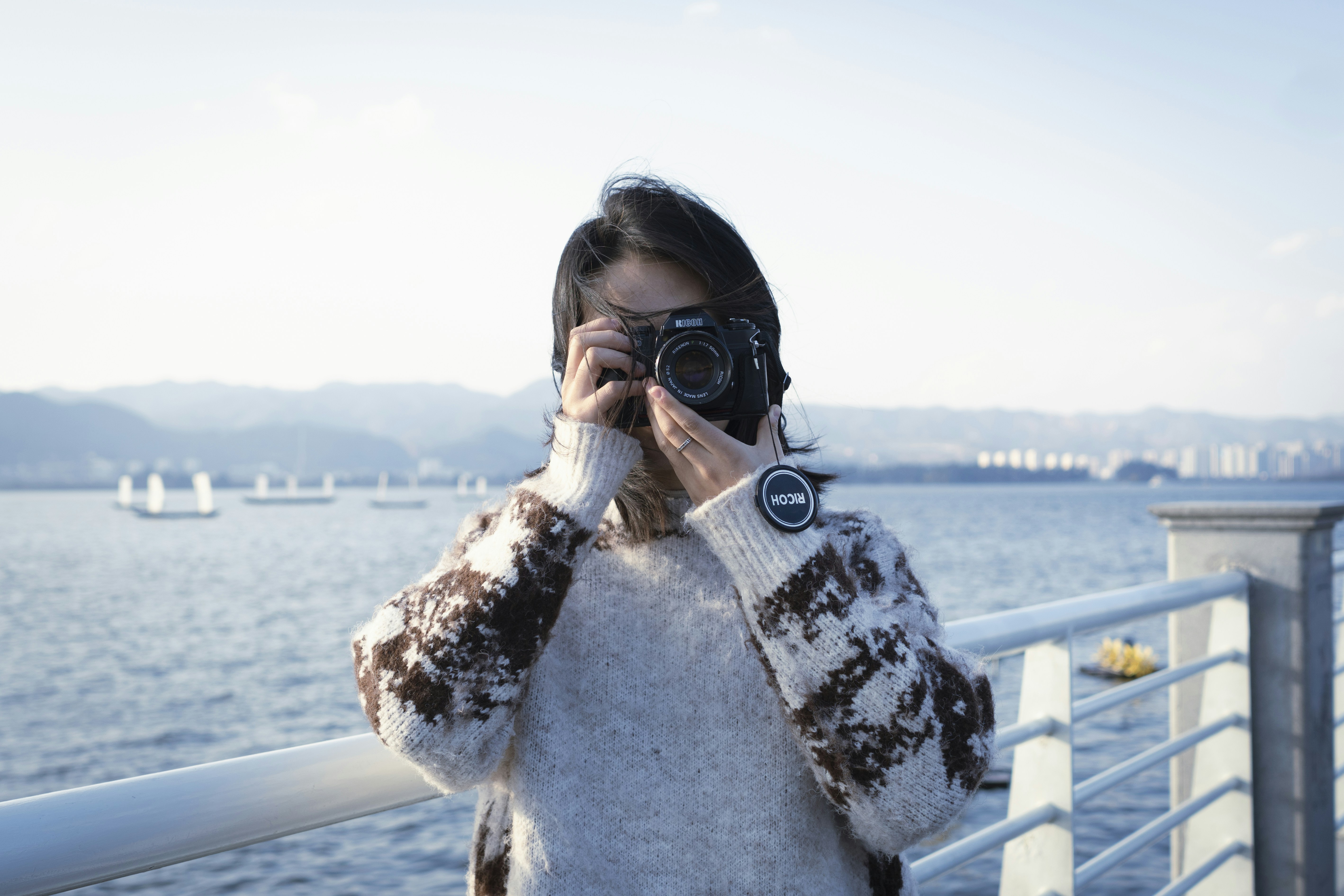 Person taking a photo on a waterfront boardwalk with sailboats in the background.