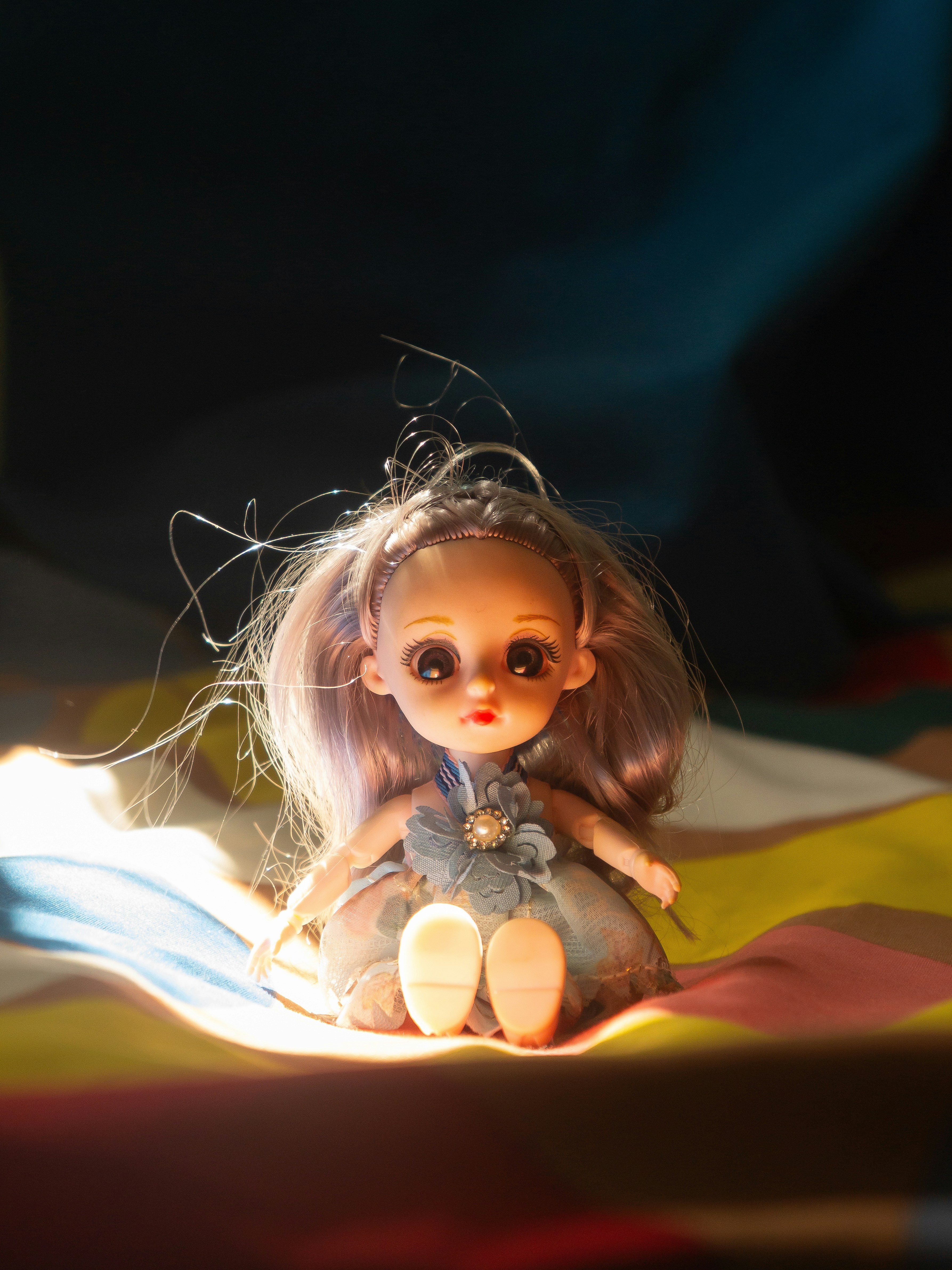 A charming doll with flowing hair sits on a colorful striped fabric, illuminated by soft sunlight that highlights its features.