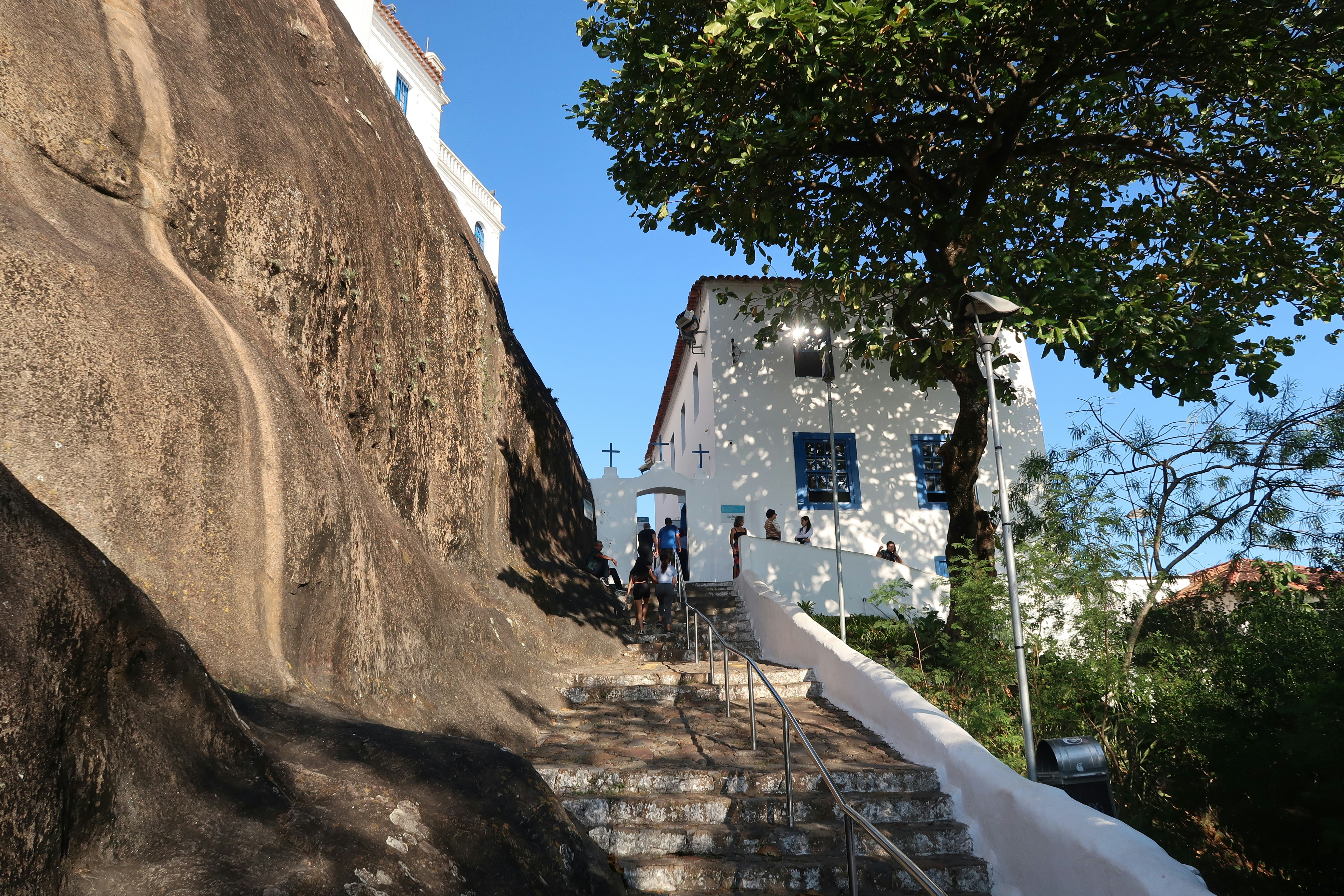 A group of people walking up a stone stairway beside a rocky cliff under a clear blue sky.