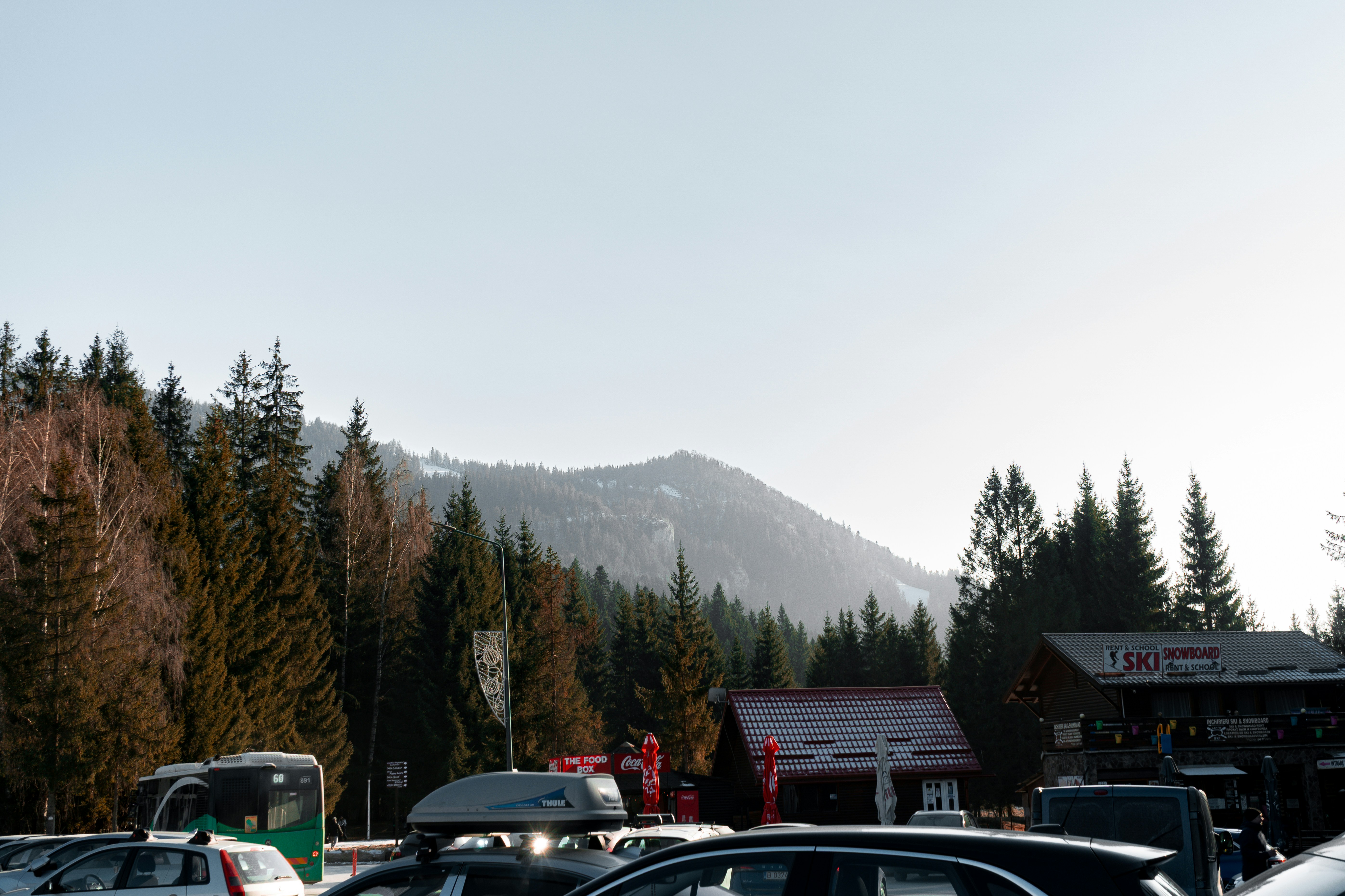 Cars parked near a ski resort with a mountain range and dense forest in the background under a clear sky.