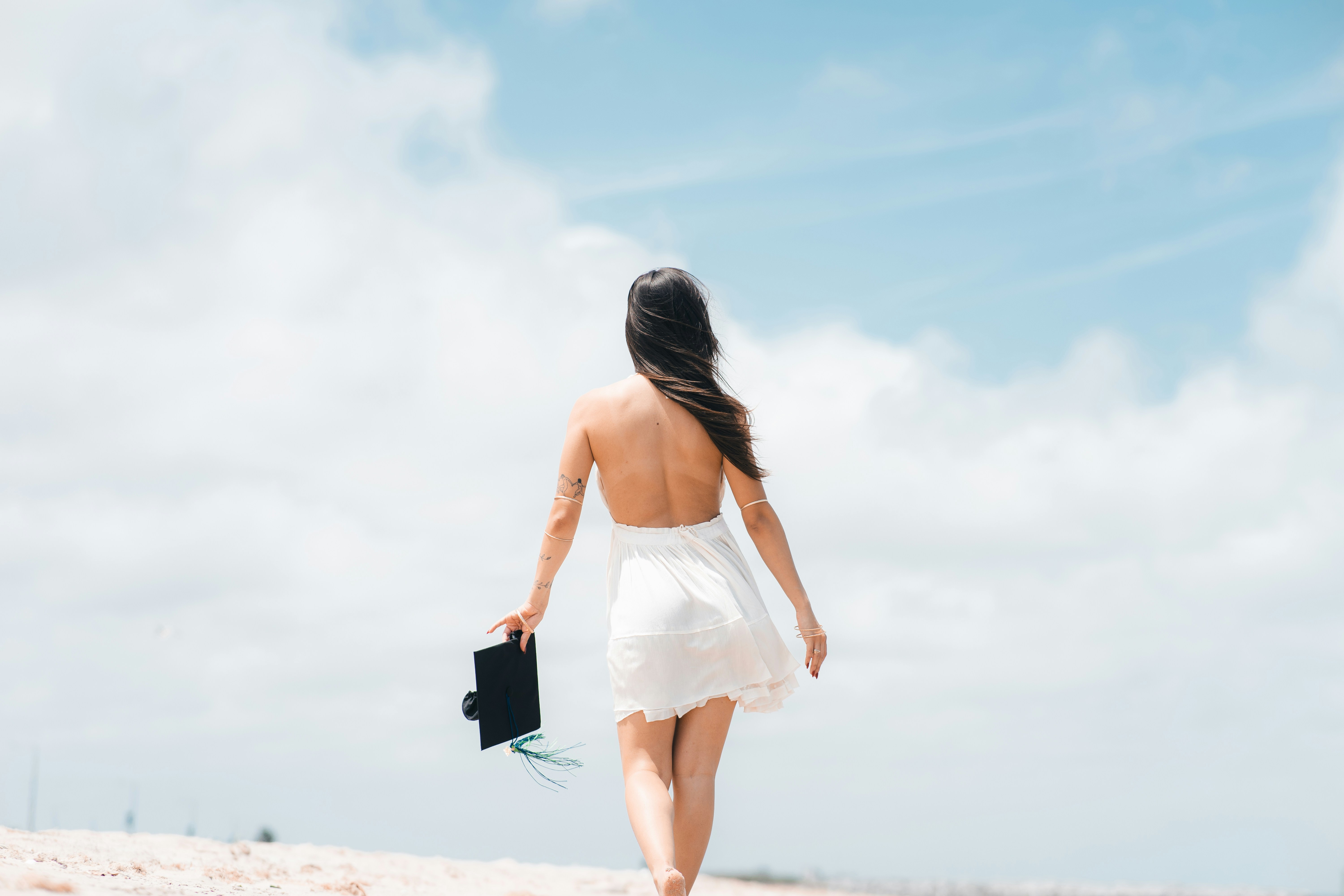 Woman in a white dress walking barefoot on a sandy beach under a bright blue sky.