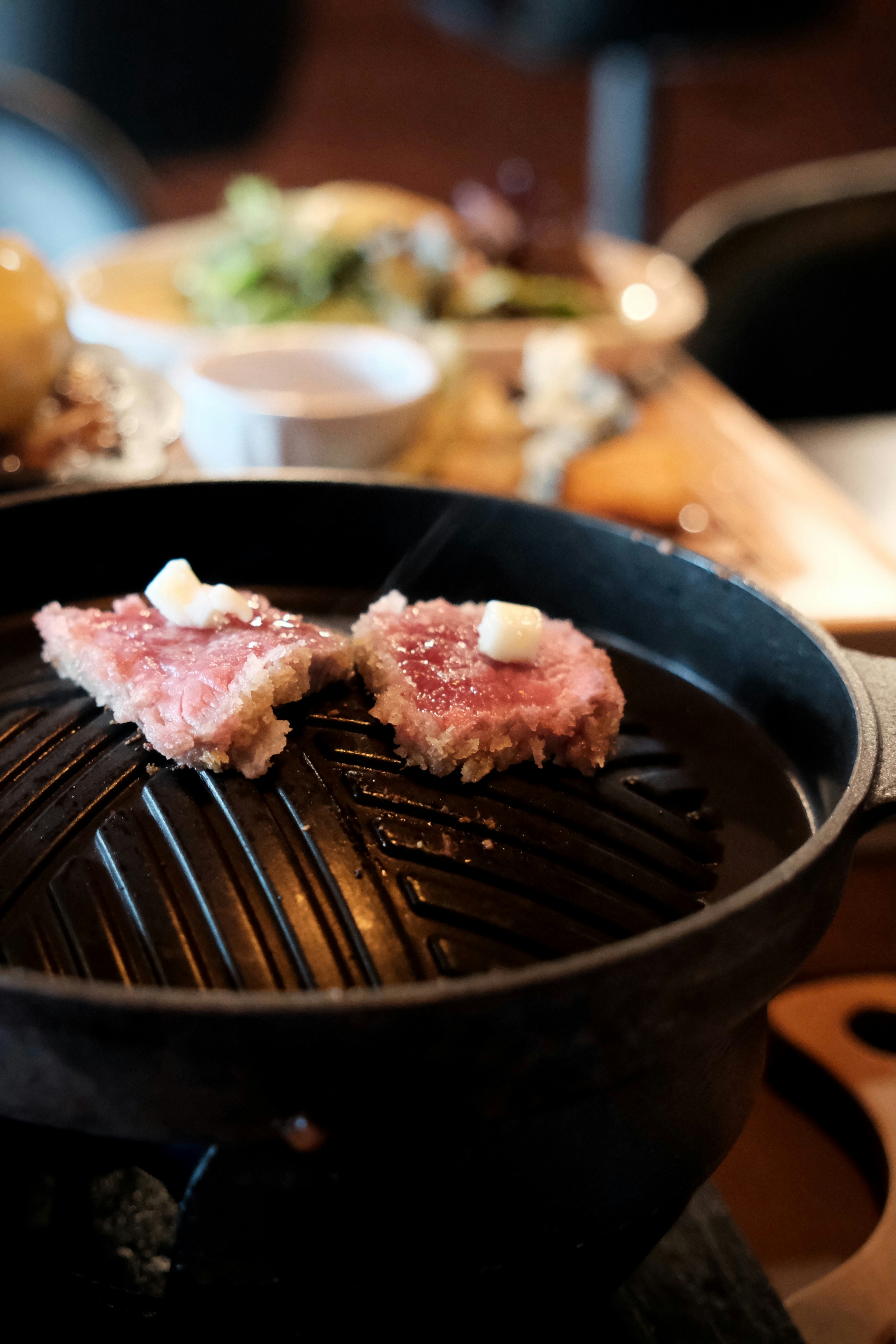 Two slices of marbled beef sizzling on a grill with a pat of butter melting on top. Background features an assortment of colorful dishes.