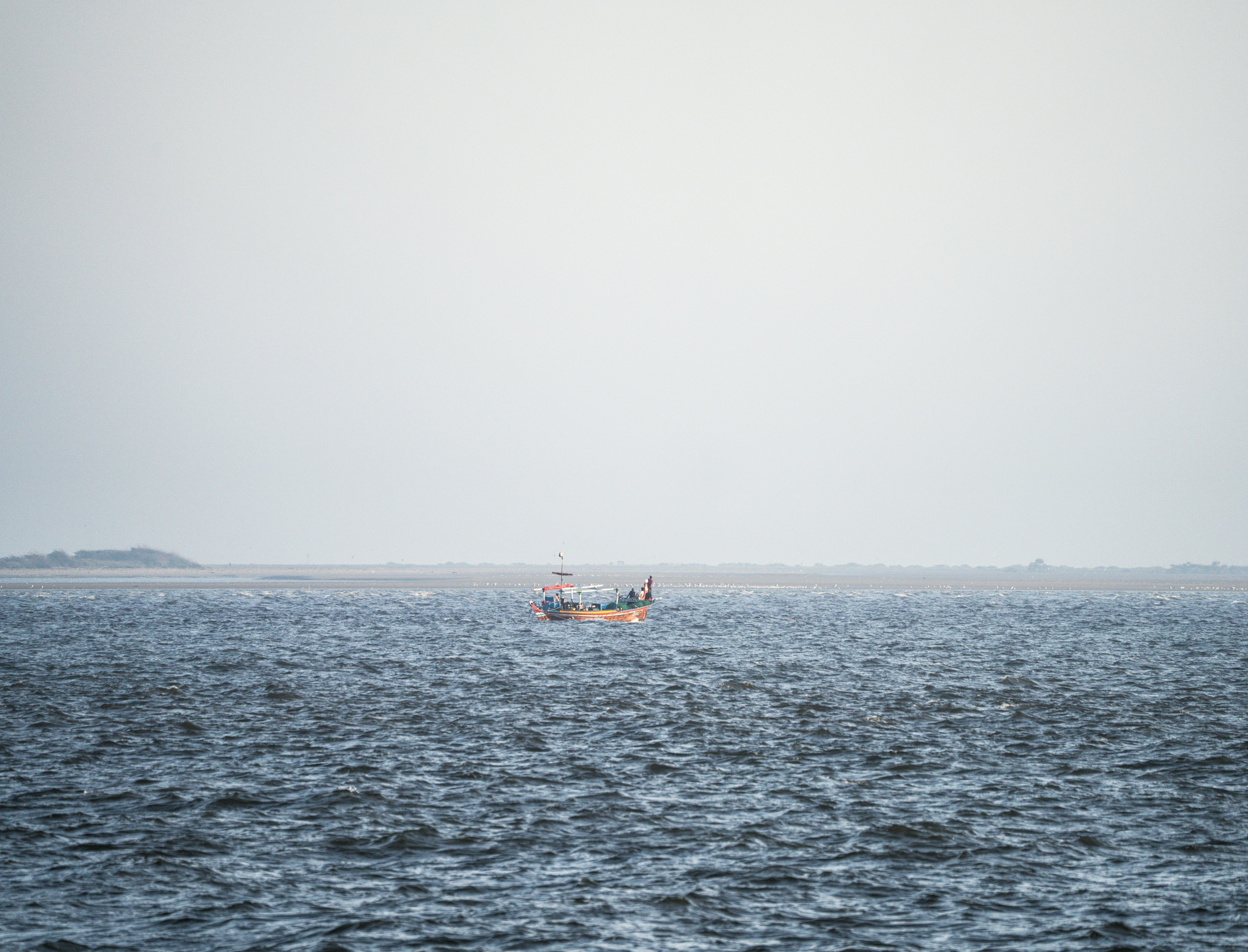 Small boat navigating vast ocean with distant shoreline under clear sky.