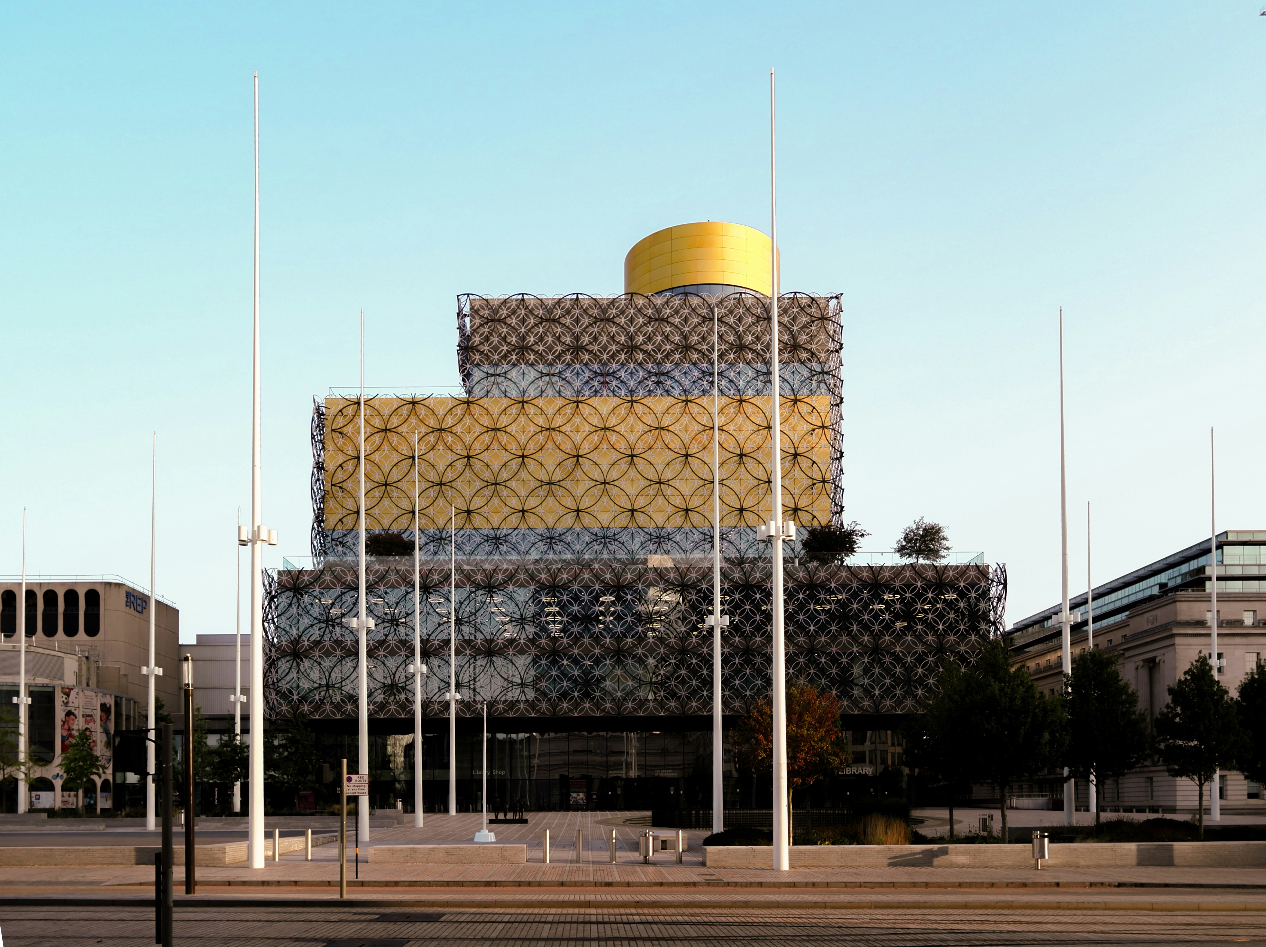 Photograph of a modern building with a lattice screen facade and a bright yellow cylindrical crown, set against a clear blue sky.