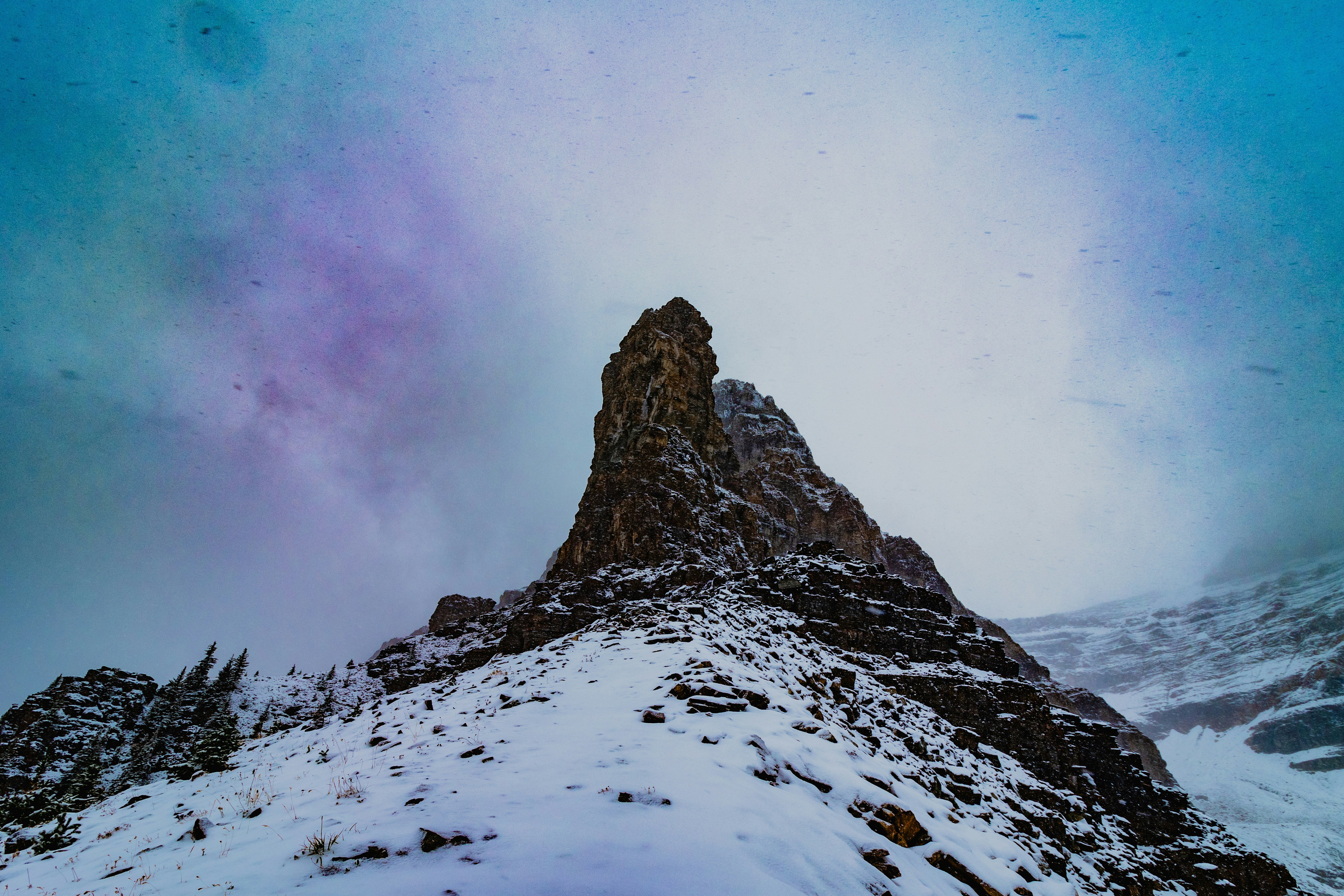 Jagged rocky peak dusted with snow beneath a swirling, colorful sky.