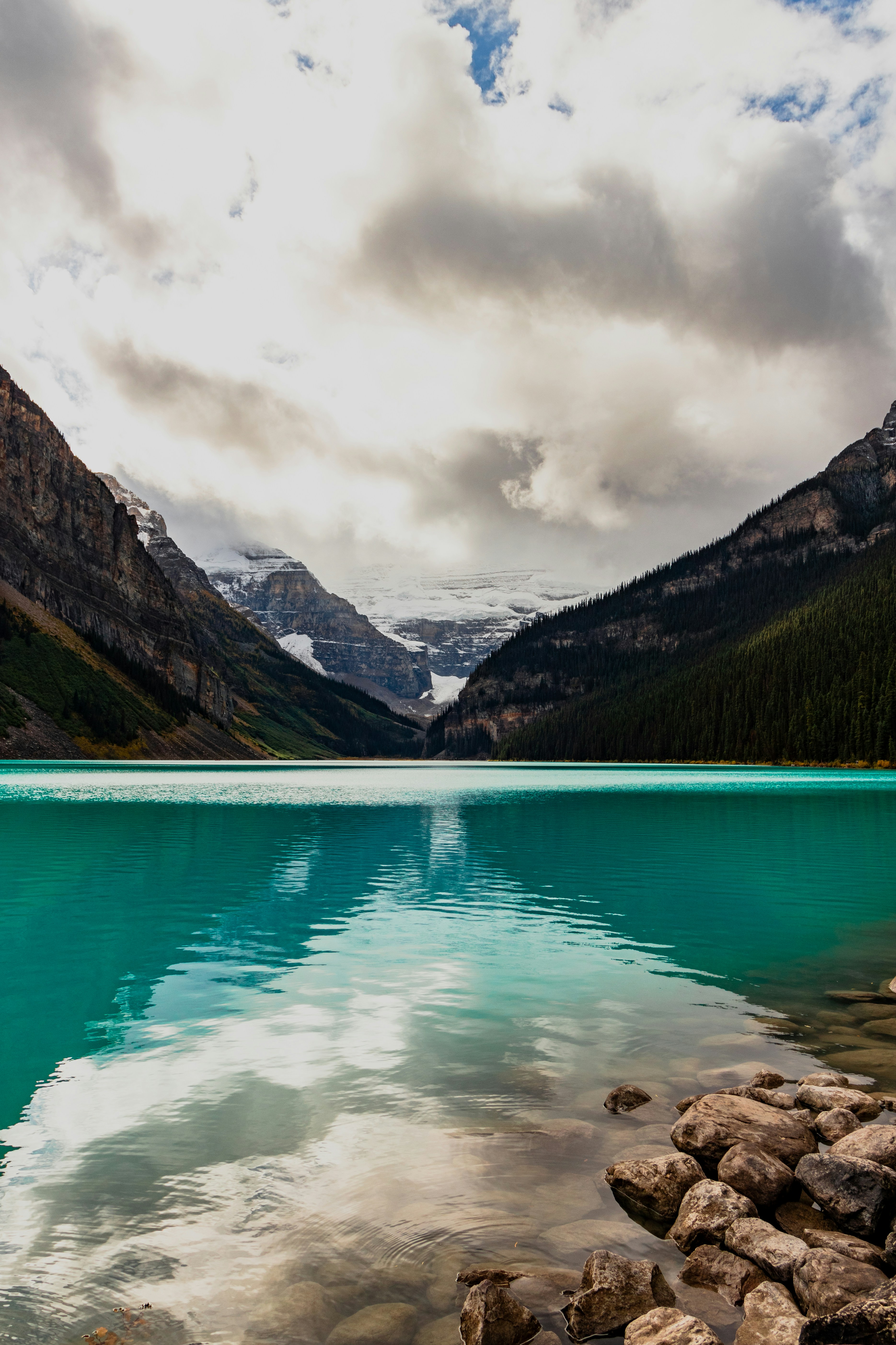 Turquoise lake reflecting majestic mountains under a cloudy sky, framed by lush forests and rocky shores.