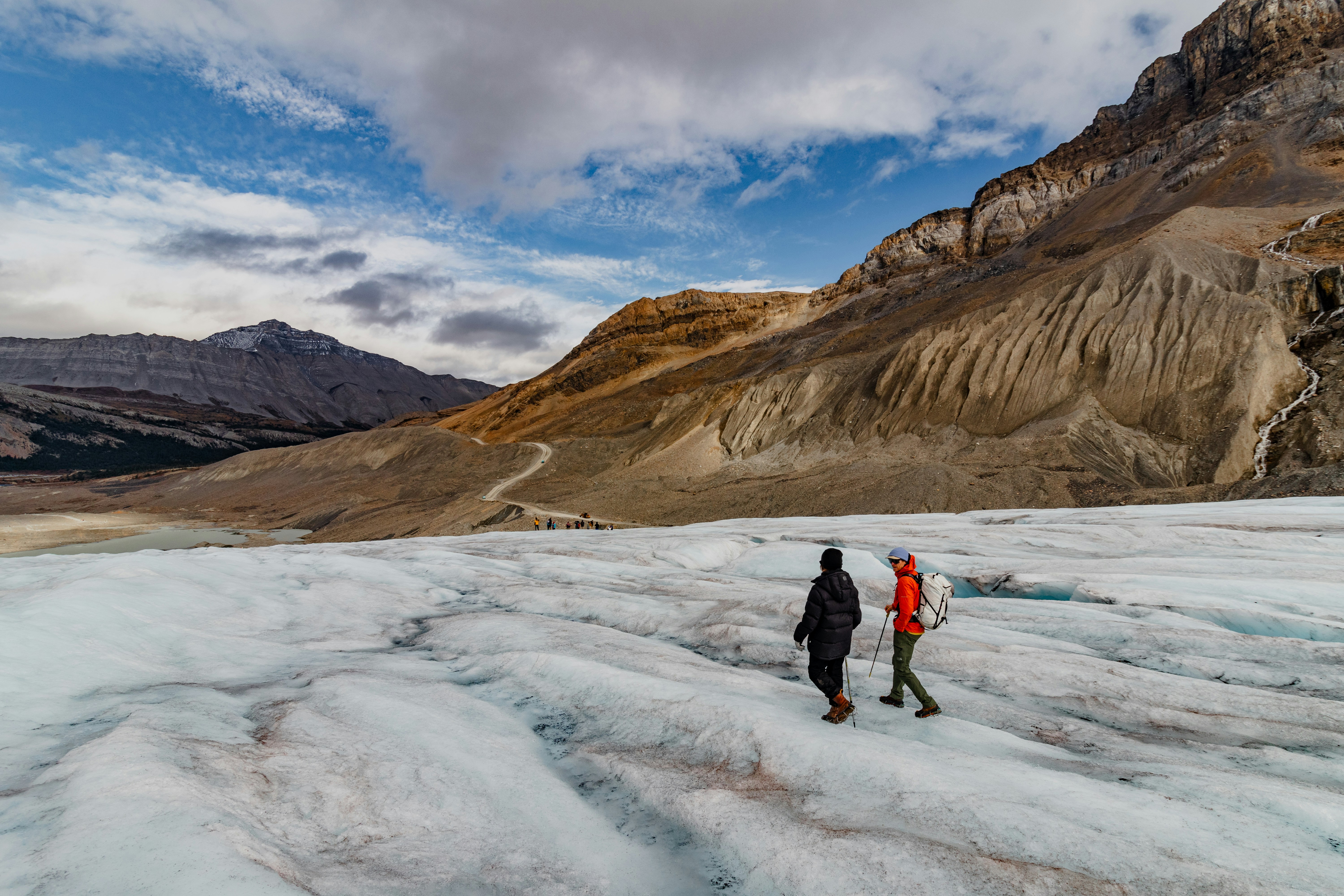 Two hikers traverse a glacier with rugged mountains in the background under a partly cloudy sky.