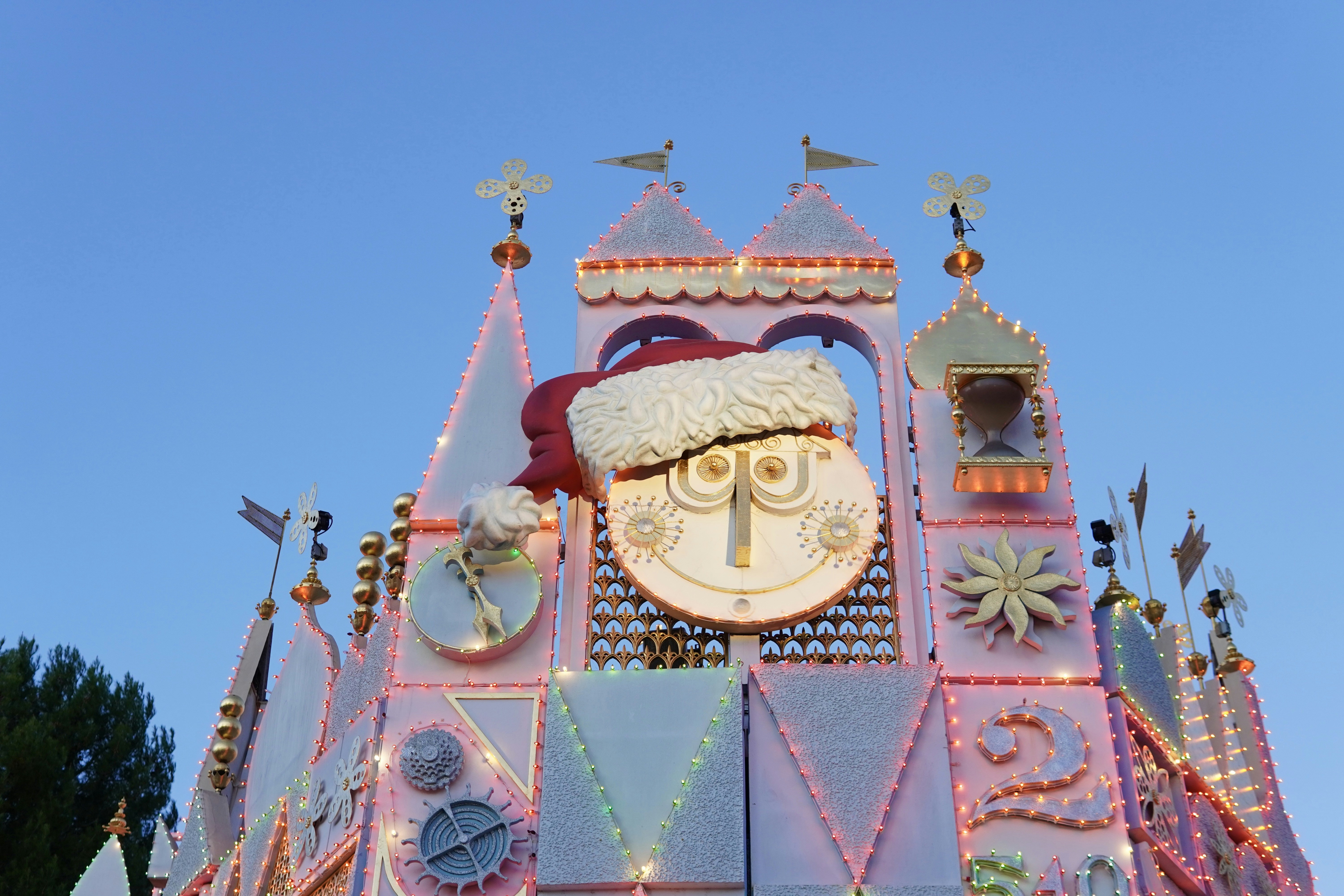 Festive facade of 'it's a small world' adorned with colorful lights and Santa hat under a clear blue sky.
