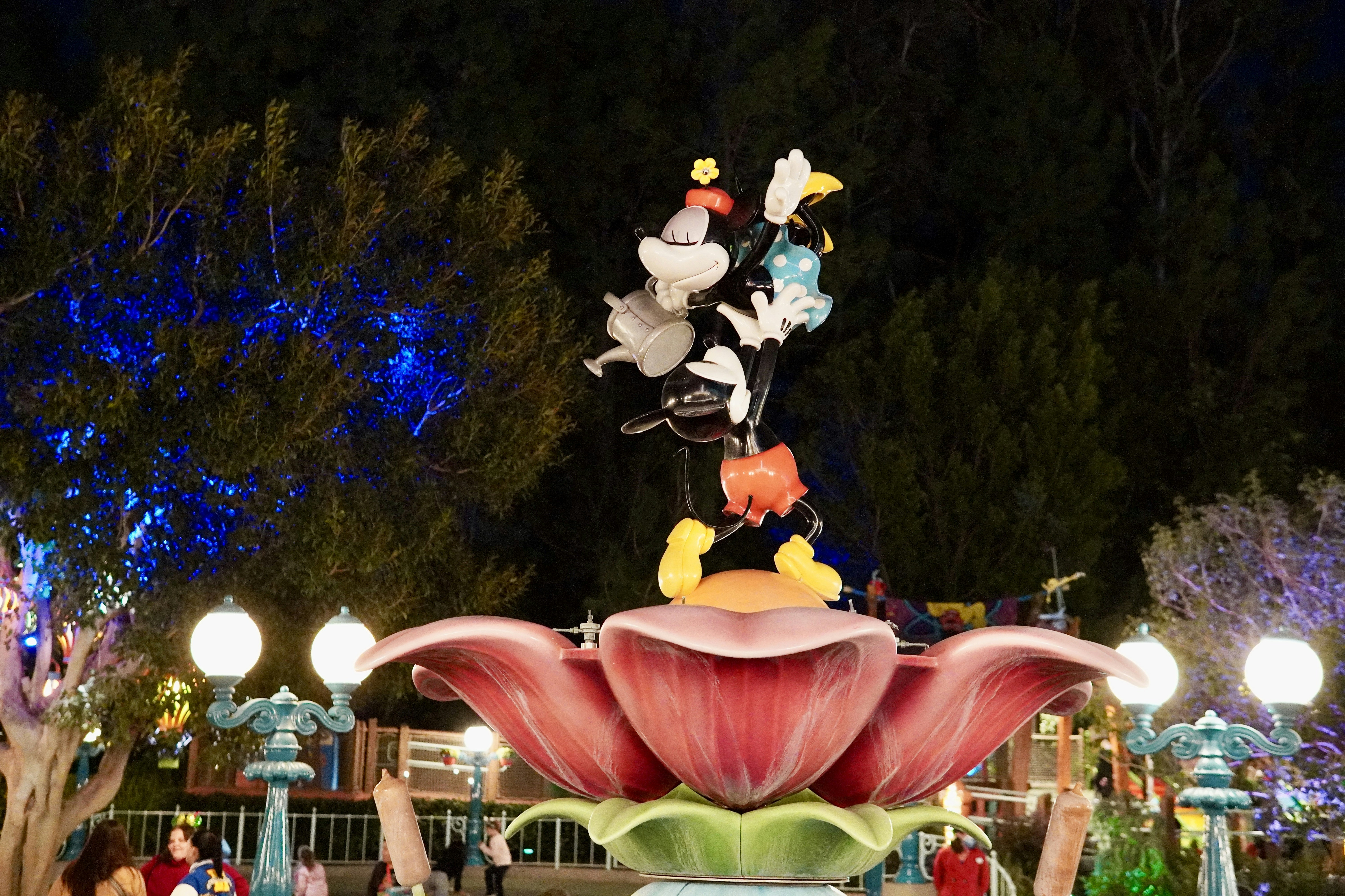Mickey and Minnie atop a giant flower fountain, illuminated by colorful lights in Disneyland's Toontown at night.