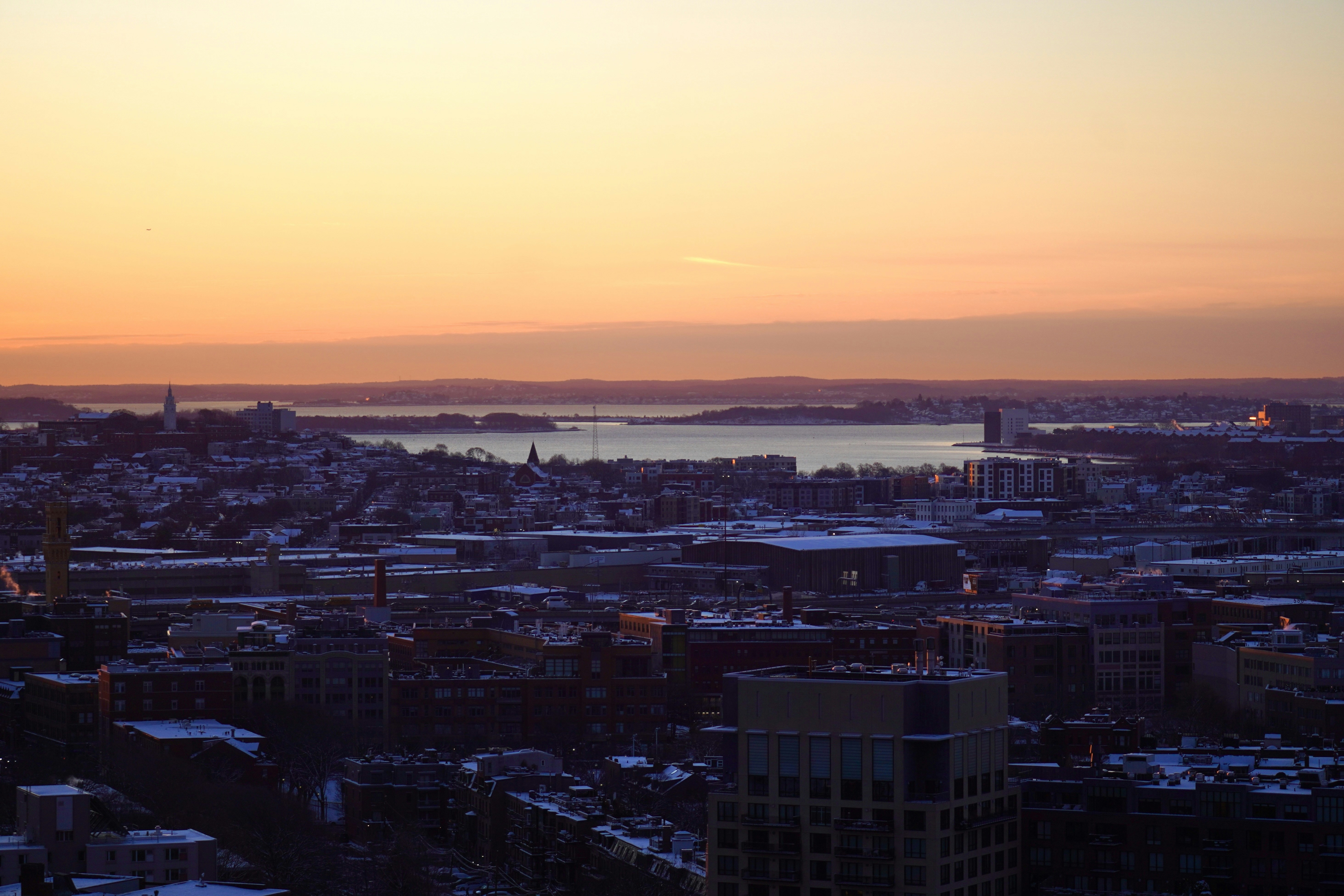 Boston skyline at sunrise with snow-covered rooftops and warm orange sky over Boston Harbor.