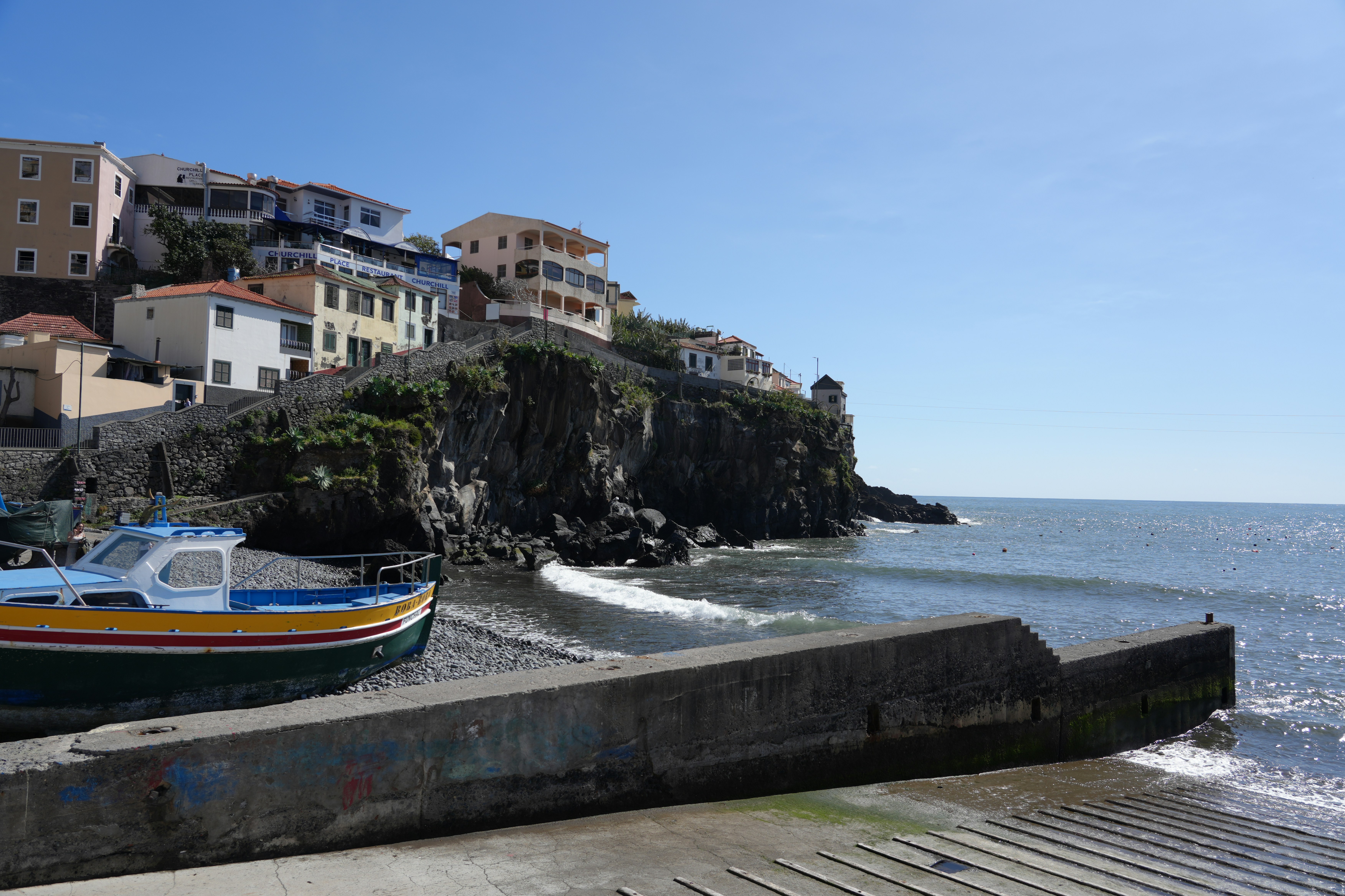 Colorful fishing boats rest by a rocky cliffside in Câmara de Lobos, Madeira, under a clear blue sky.