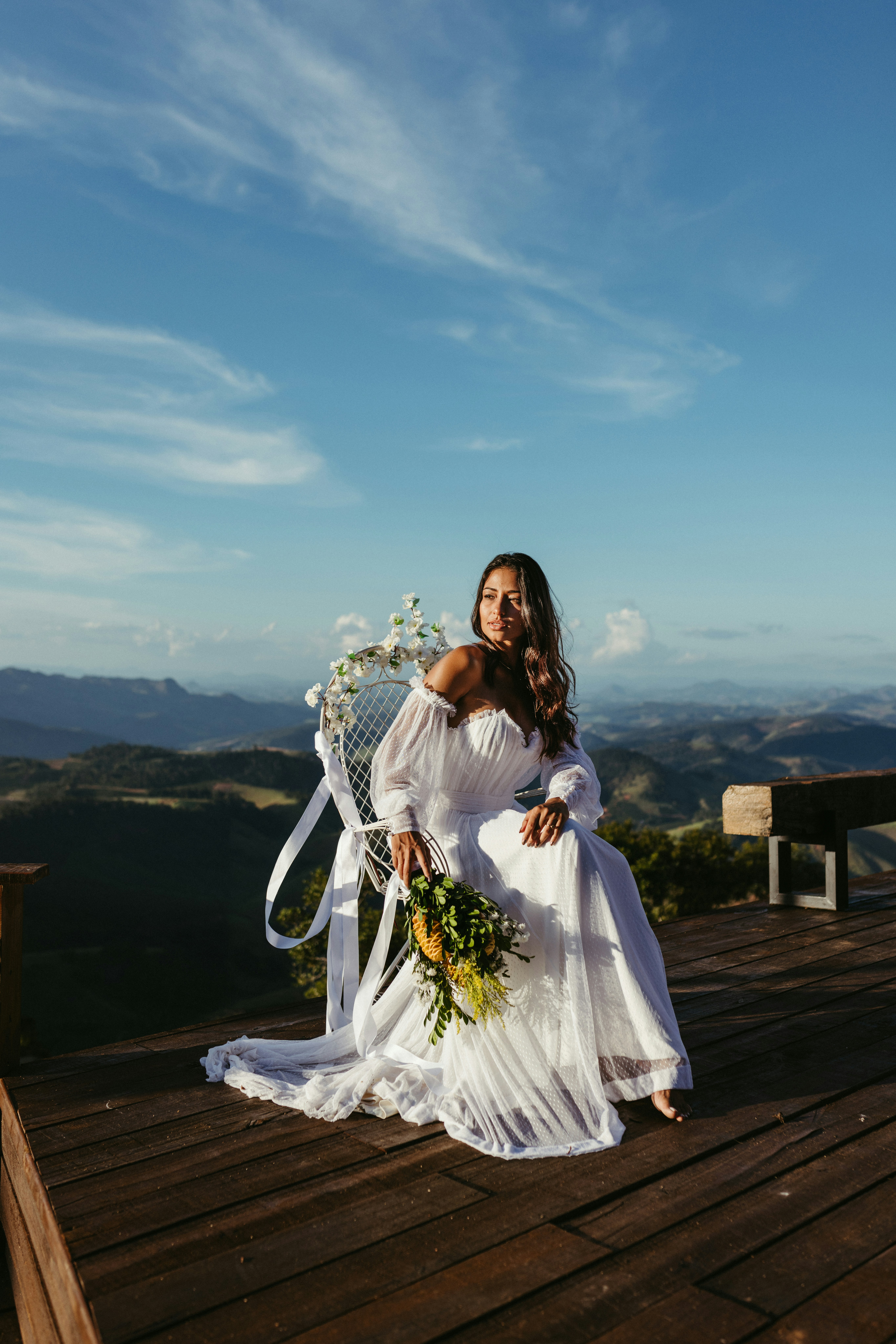 Hillside vows on a weathered deck: bride in white with bouquet against a sweeping mountain backdrop