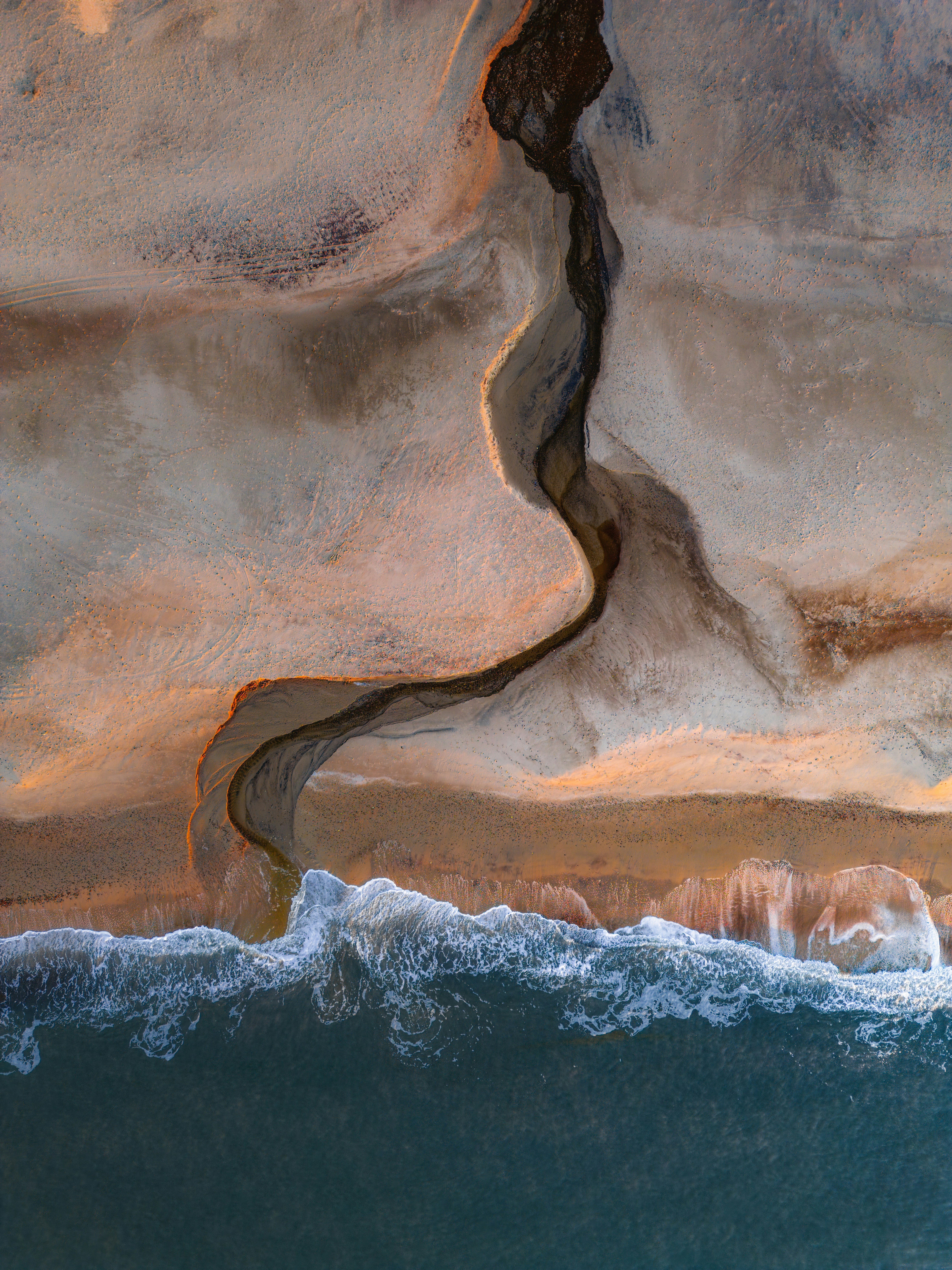 Aerial view of a rugged coastline with waves meeting a sandy shore, dissected by a winding crevice.