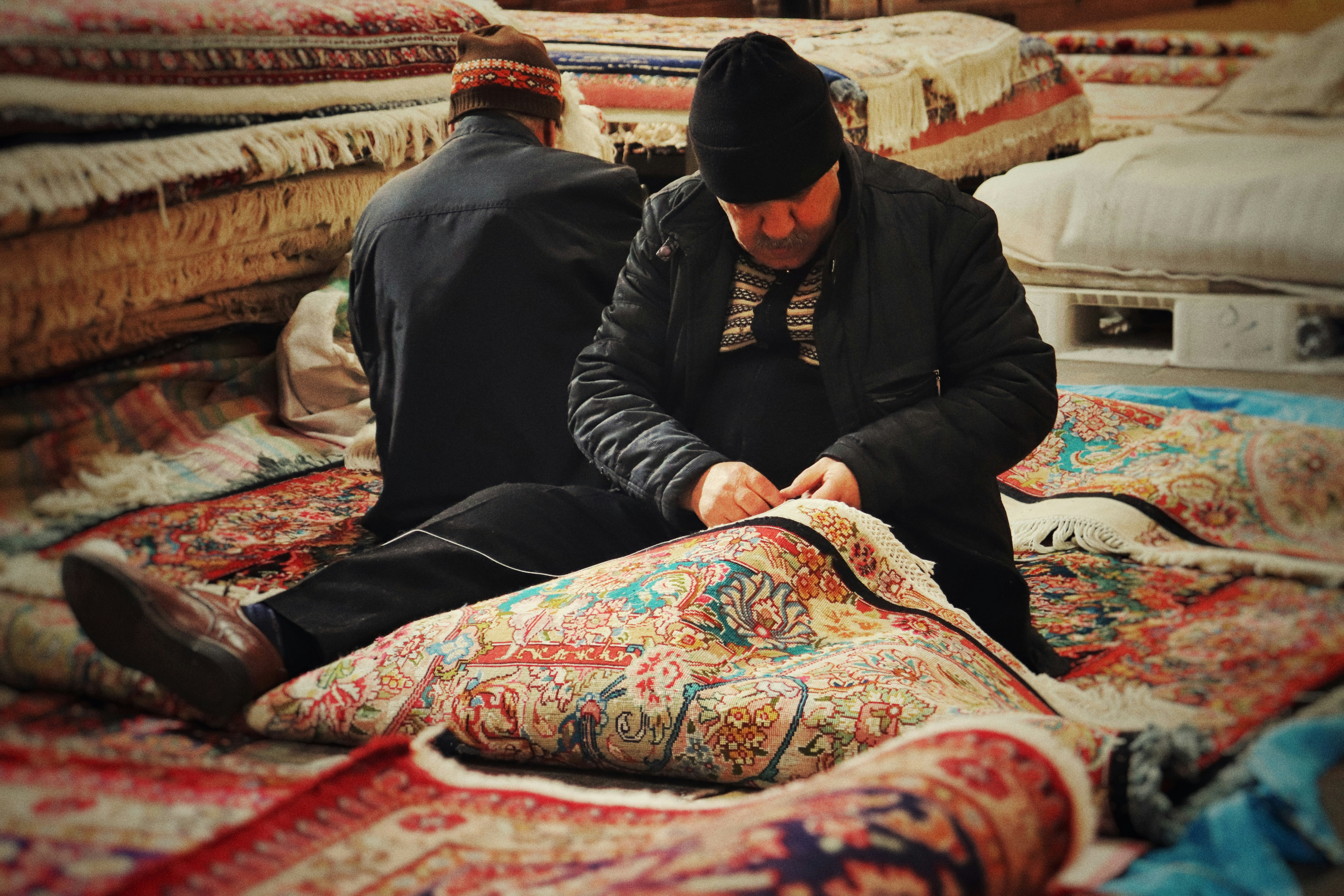 Man sewing ornate patterns into a colorful rug, surrounded by stacked textiles.