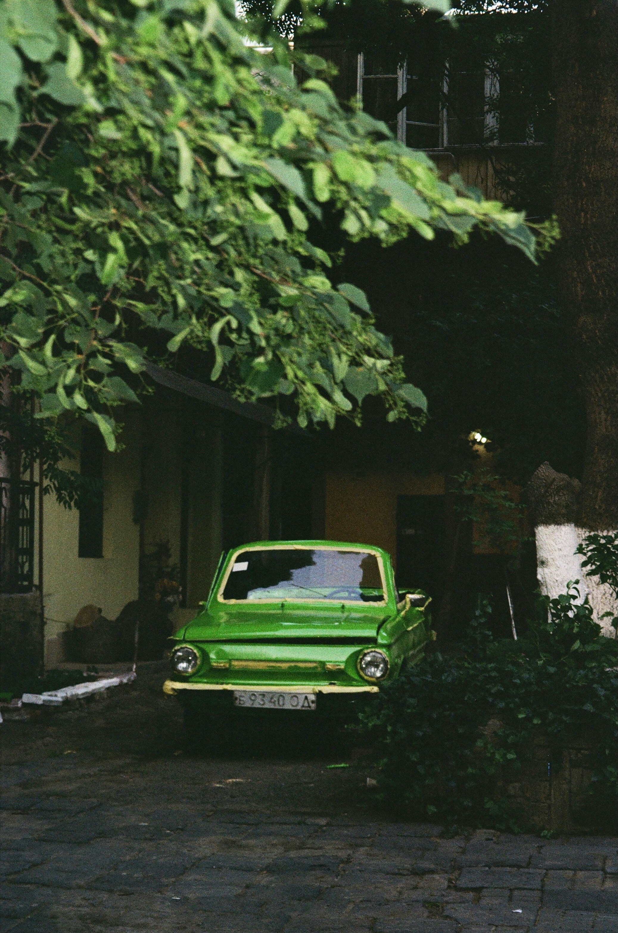 A bright green vintage car sits parked in a shaded courtyard, framed by ivy and a dark doorway behind.