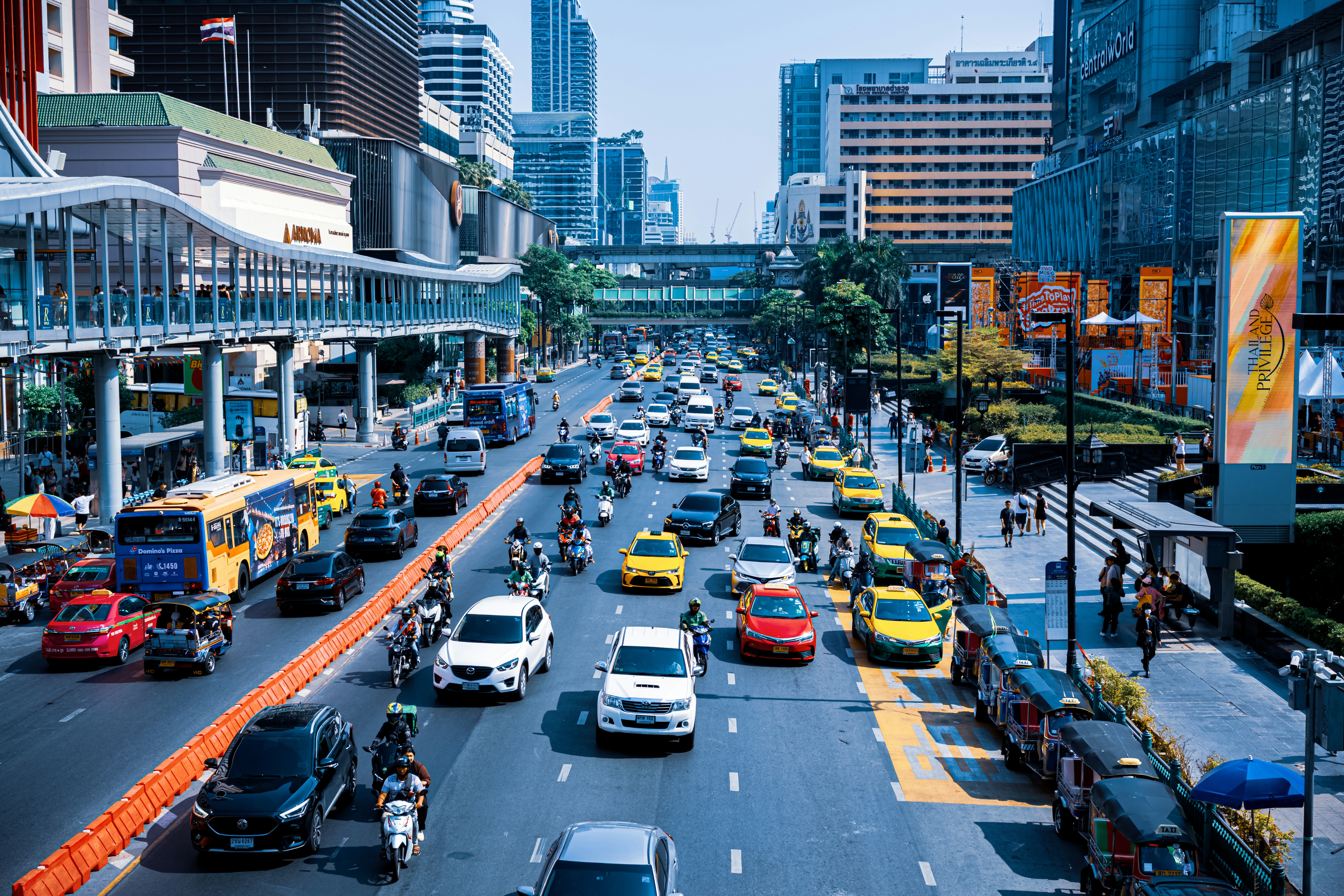 Busy Bangkok street filled with colorful taxis and motorbikes under towering skyscrapers on a sunny day.