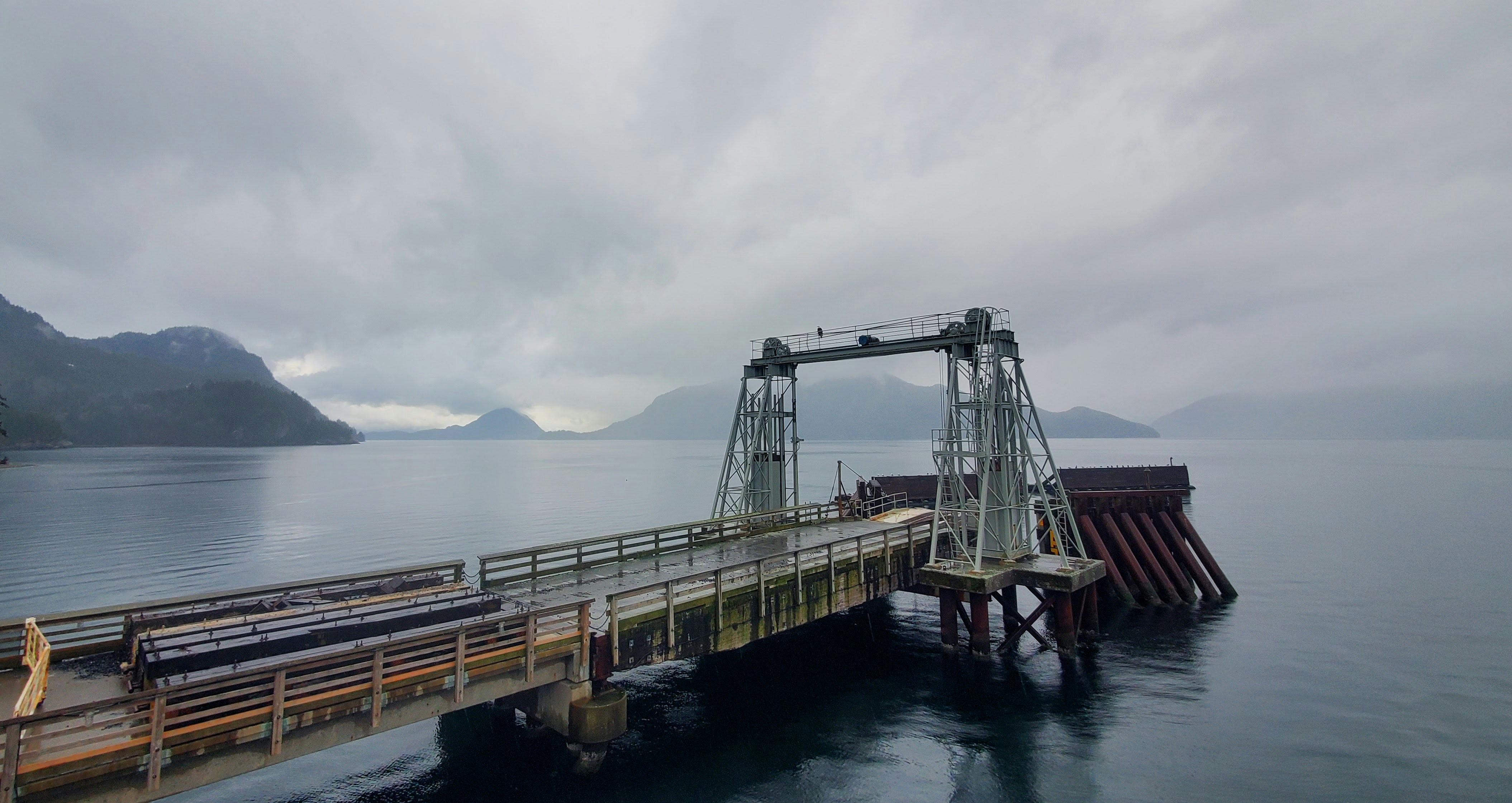 Overcast sky above a solitary dock extending into a calm lake with misty mountains in the background.