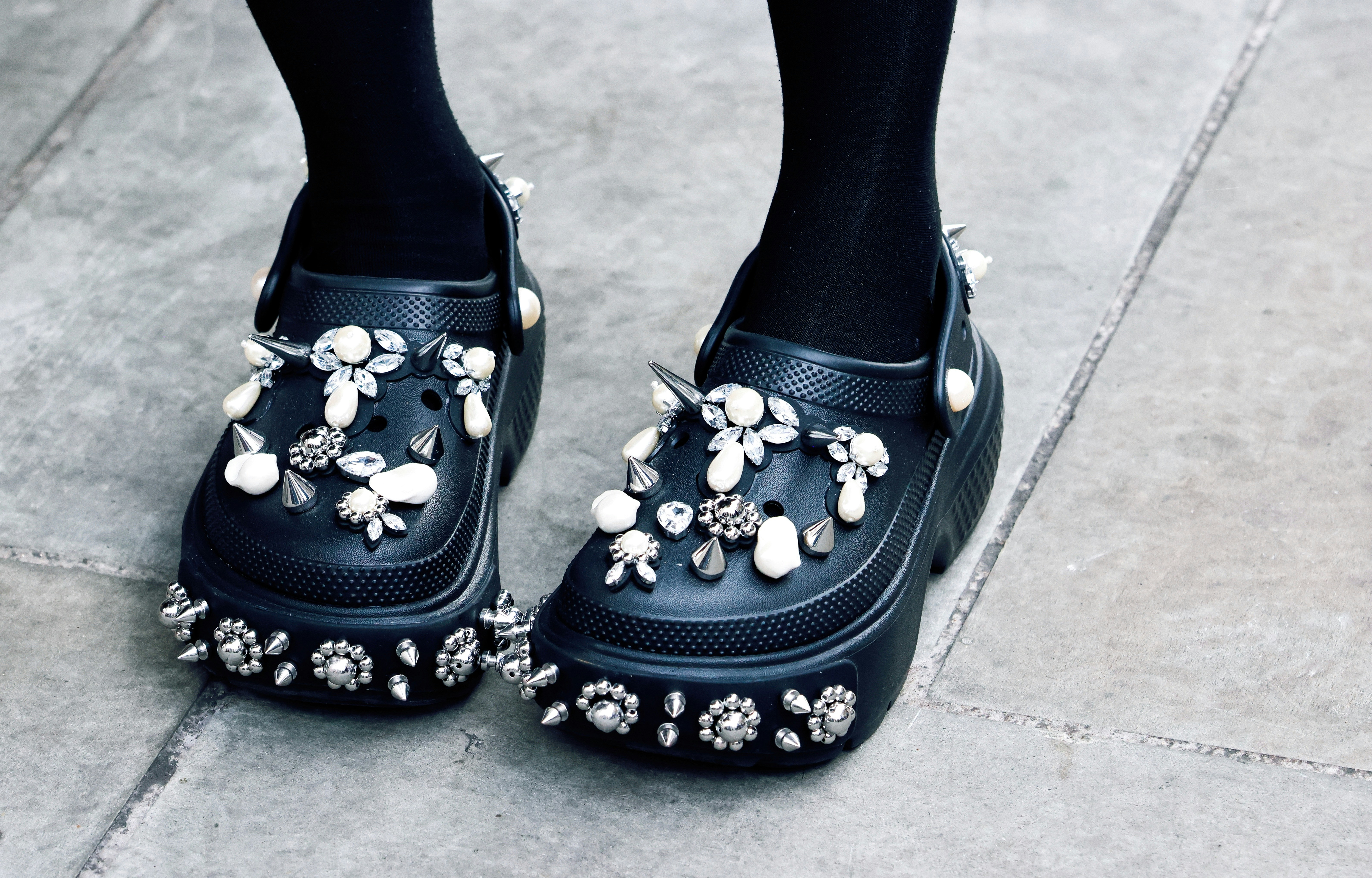 Black platform clogs adorned with silver and pearl embellishments on a stone sidewalk.